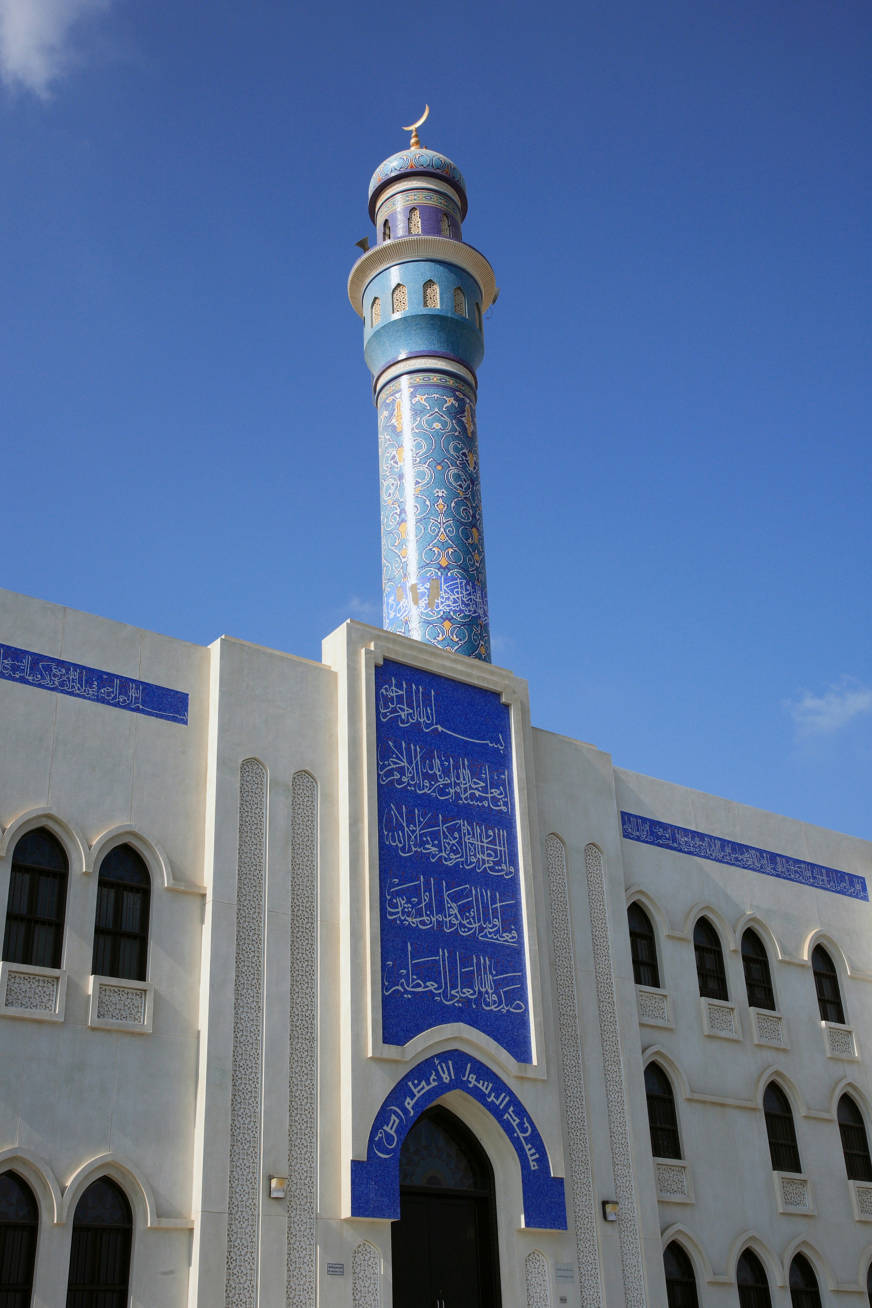 A beautifully designed mosque featuring intricate blue tile work and Arabic calligraphy against a bright sky. The minaret stands tall, showcasing the architectural splendor.