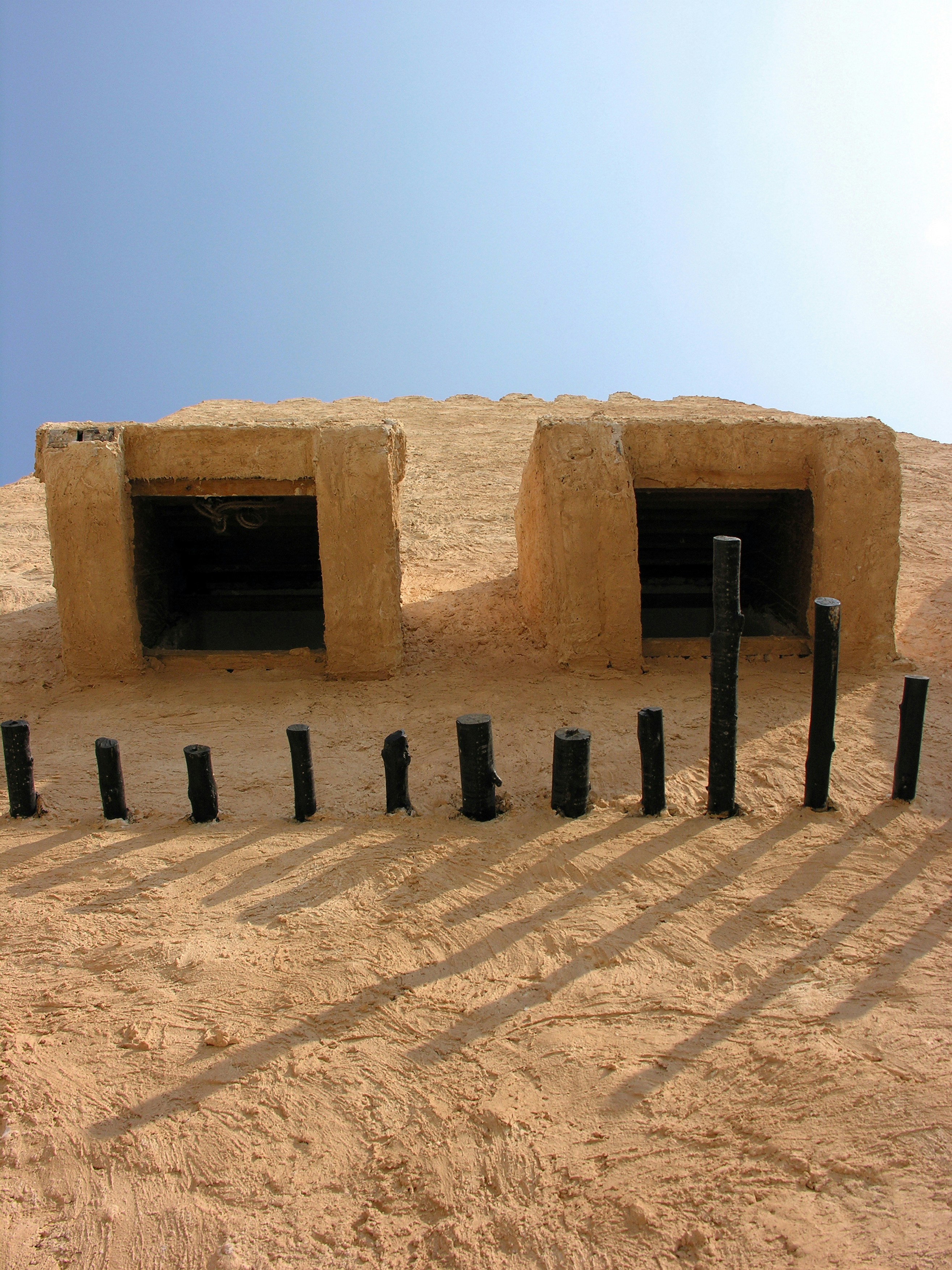 Two square windows framed by textured walls, with shadows cast by protruding wooden beams. The clear sky serves as a backdrop.