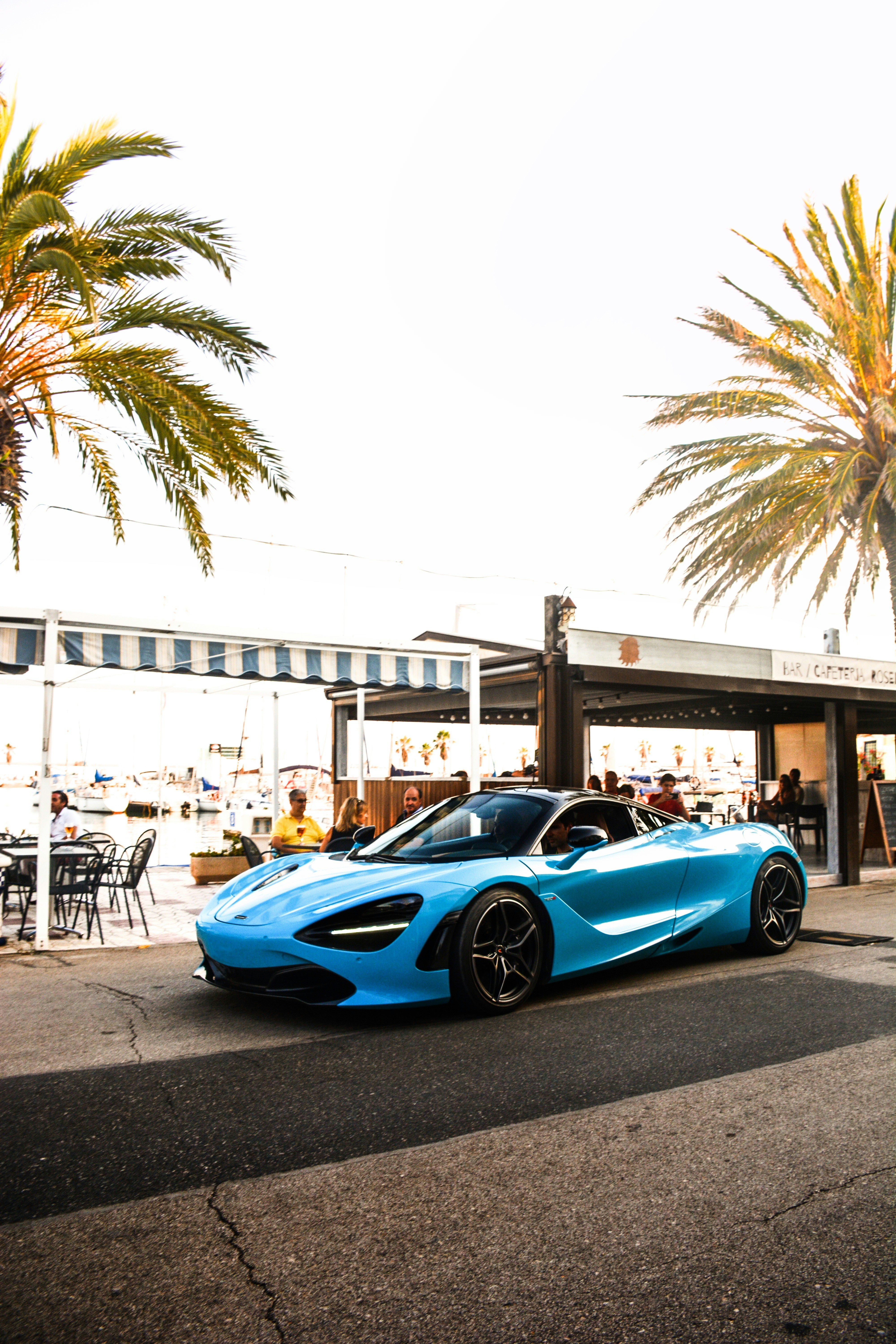 Sleek blue sports car parked beside palm trees and a bustling waterfront restaurant, embodying luxury and leisure.