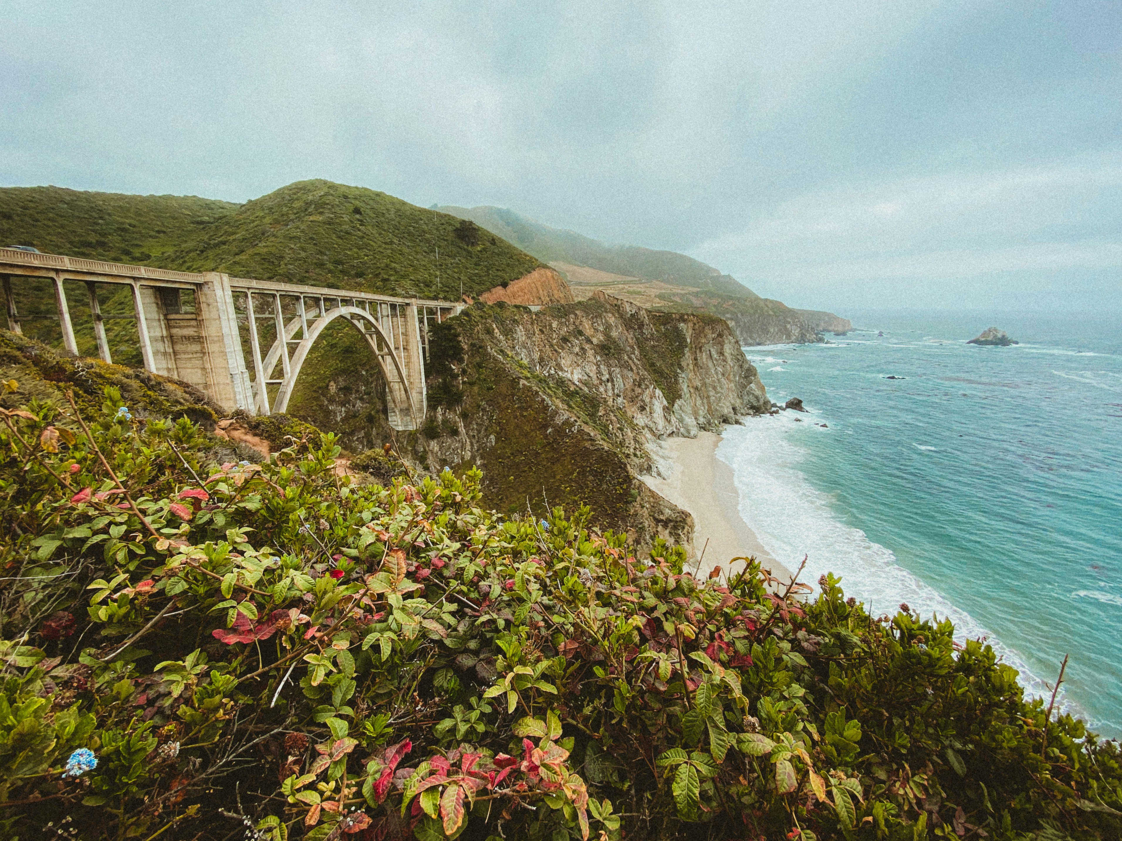 Historic arched bridge overlooking a rugged coastline with lush greenery and gentle waves below.