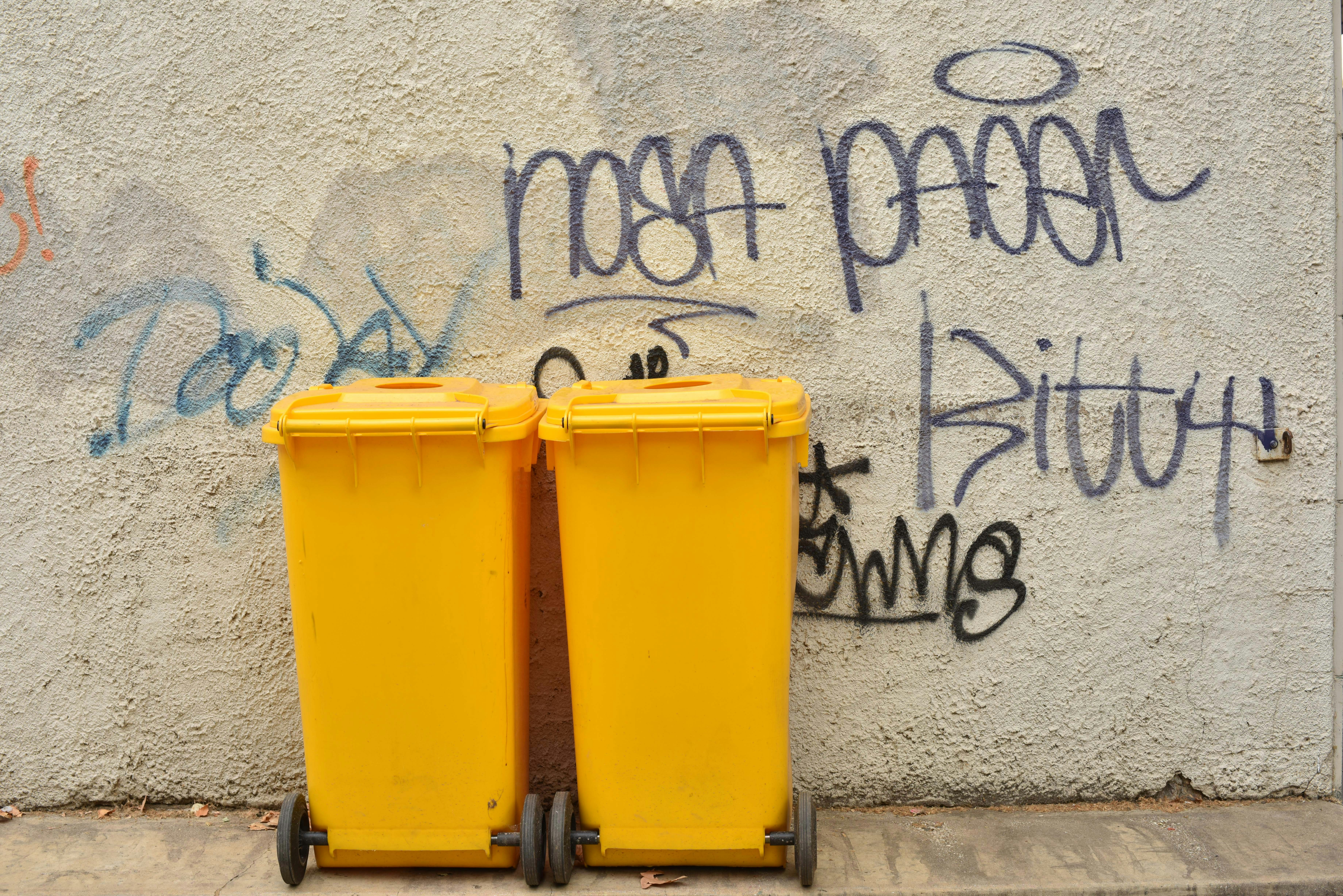 Two vibrant yellow trash bins positioned against a textured wall adorned with graffiti, showcasing urban art and everyday life.