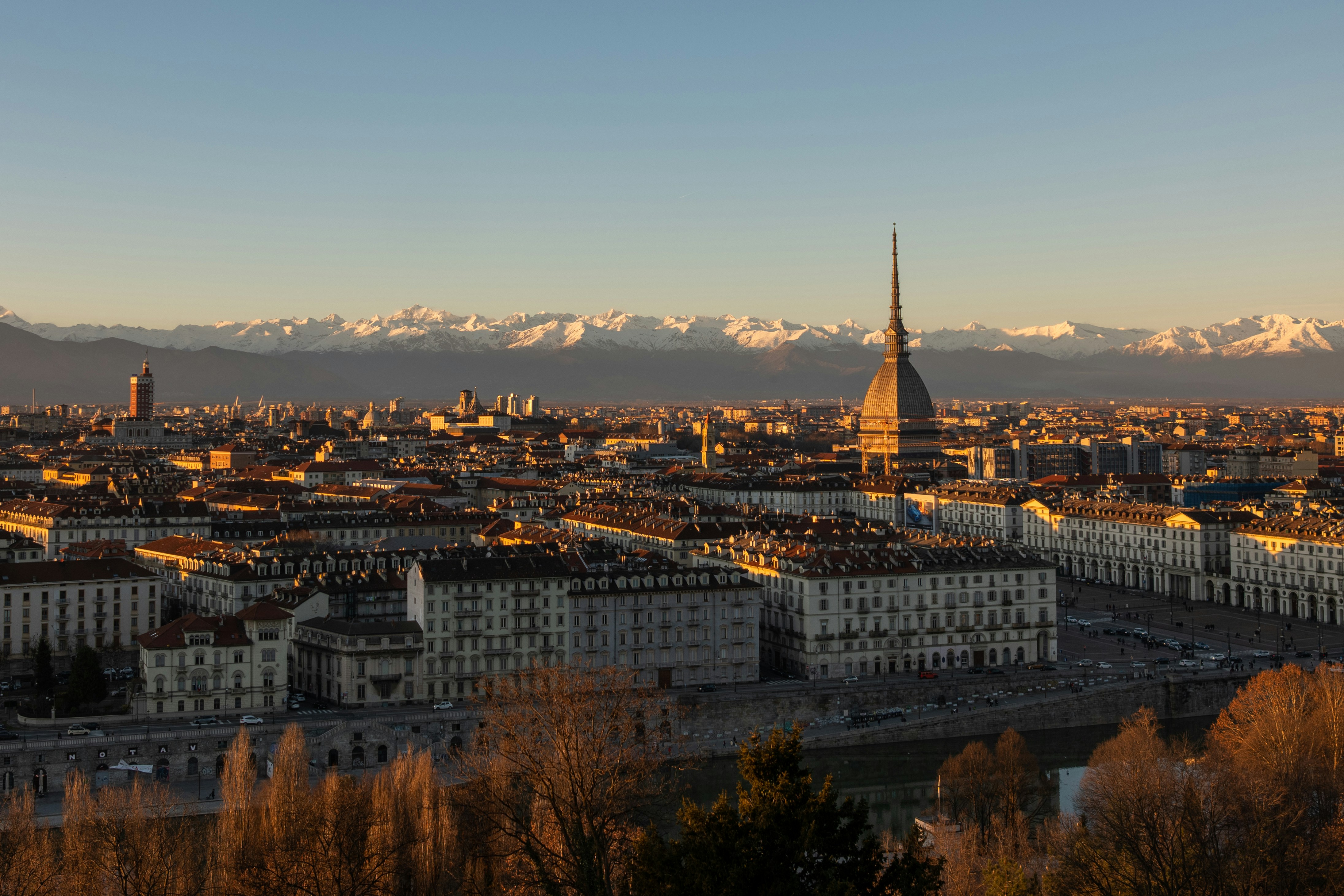 A panoramic view of Turin's cityscape at sunset, featuring the iconic Mole Antonelliana against a backdrop of snow-capped mountains.