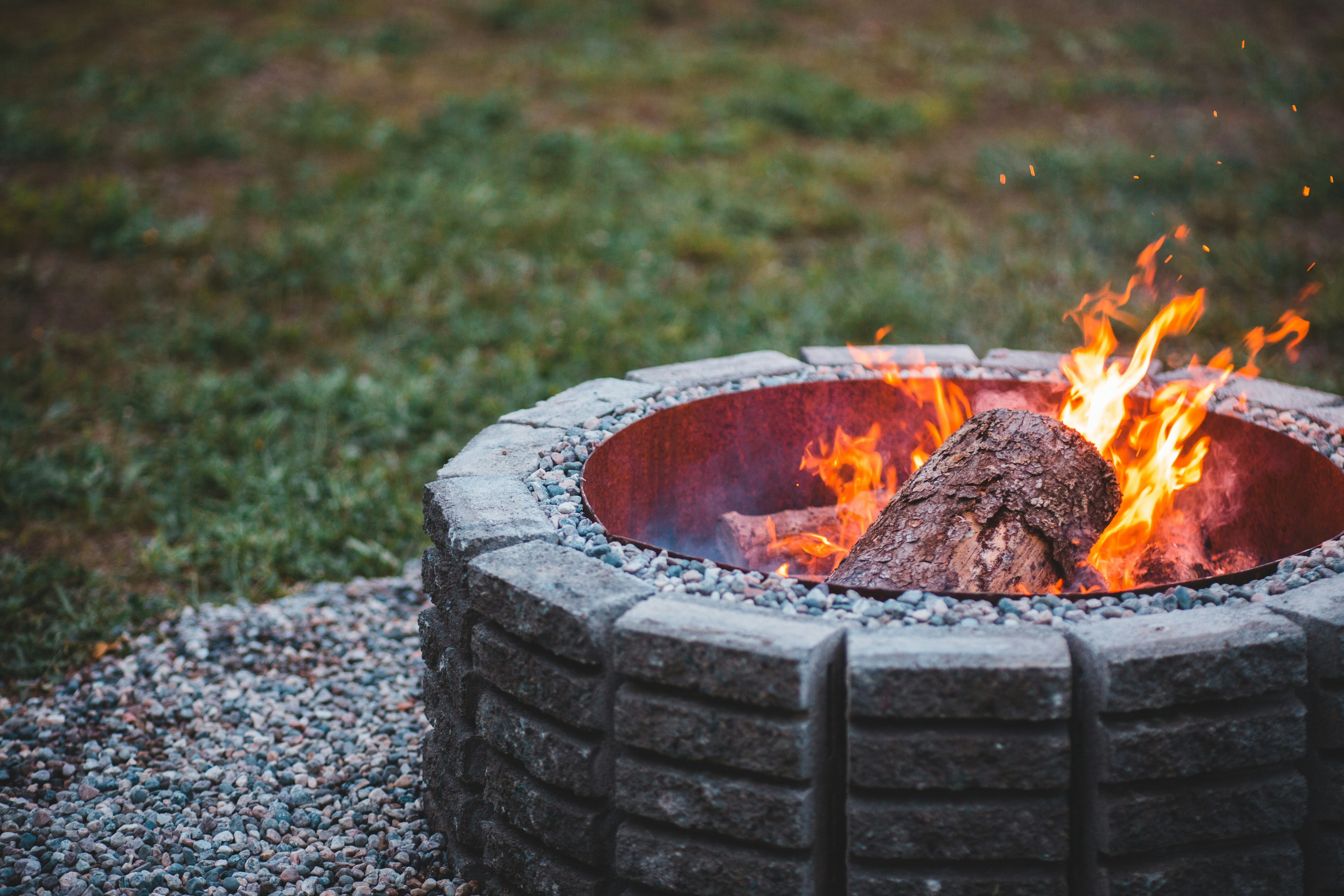 Flames dancing around a log in a circular stone firepit, surrounded by a gravel base and lush green grass. The warm glow invites a sense of community and warmth.