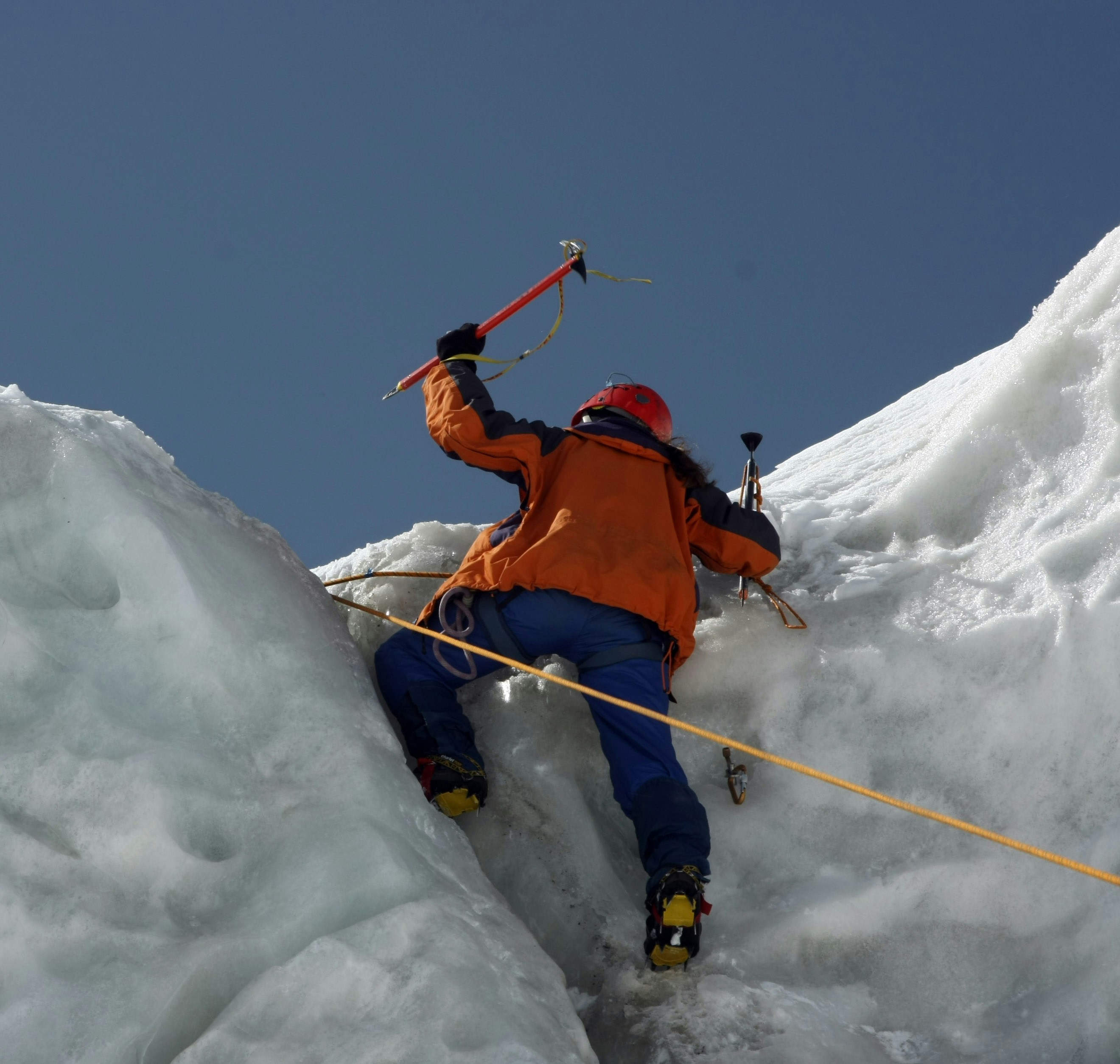 Climber navigating a steep ice formation, using tools for support and safety. Bright blue sky serves as a backdrop.