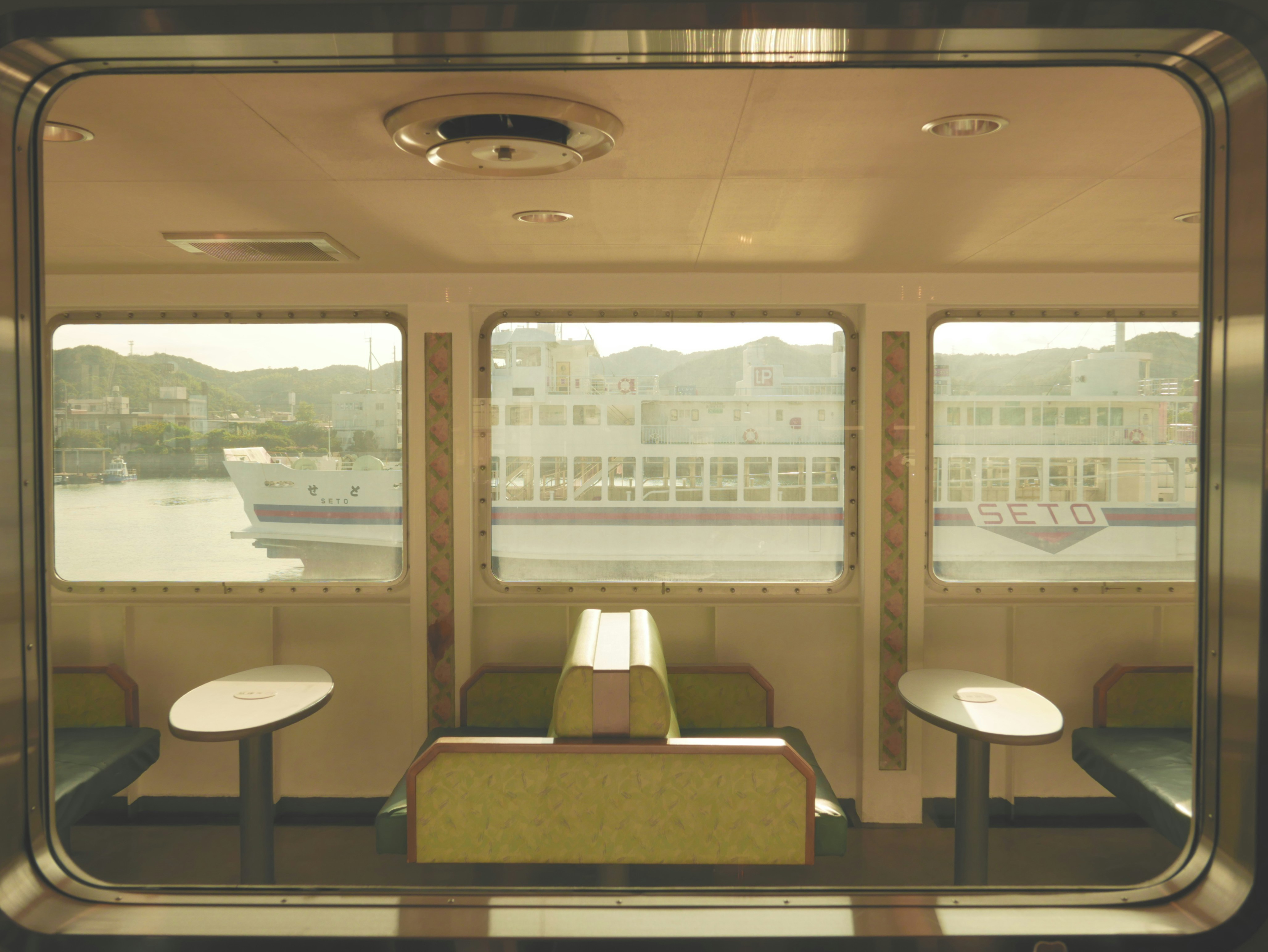 Cozy seating area inside a ferry, framed by large windows showcasing boats in the harbor. Soft light filters through, creating a calm atmosphere.