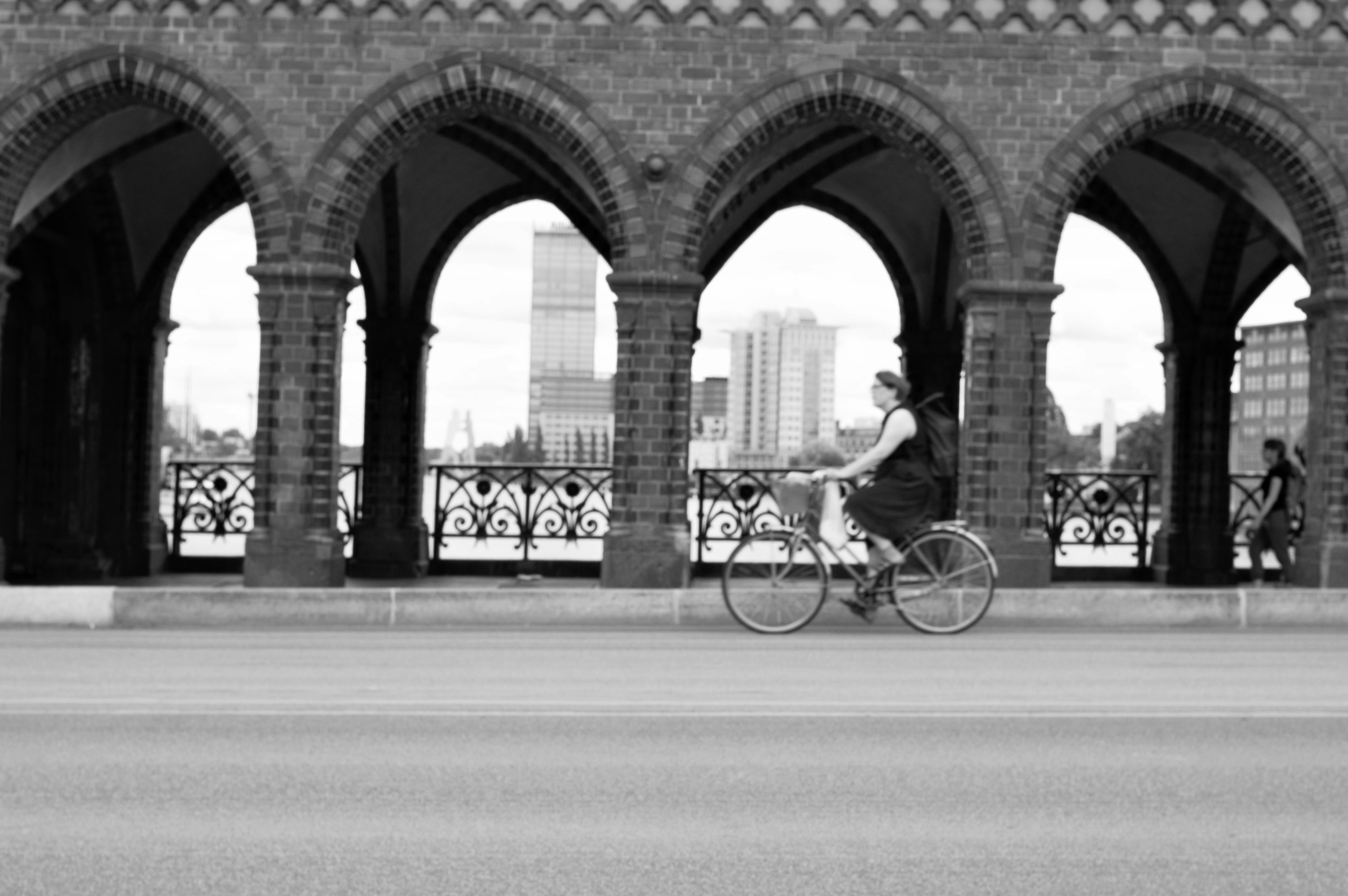 A cyclist glides past a series of ornate arches, framed by a city skyline in the background. The monochrome palette emphasizes the architectural details and motion.