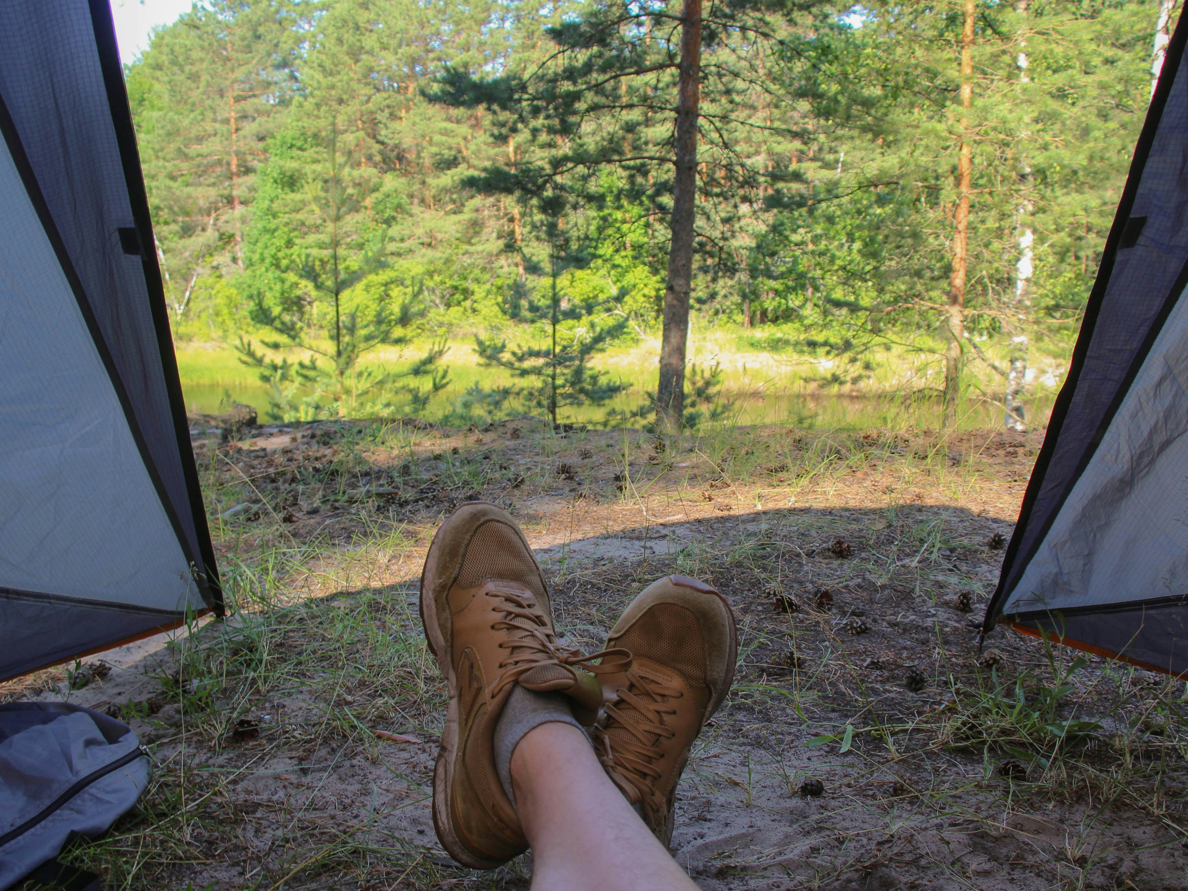 A pair of boots resting on sandy ground, framed by the entrance of a tent, revealing a lush green forest beyond.