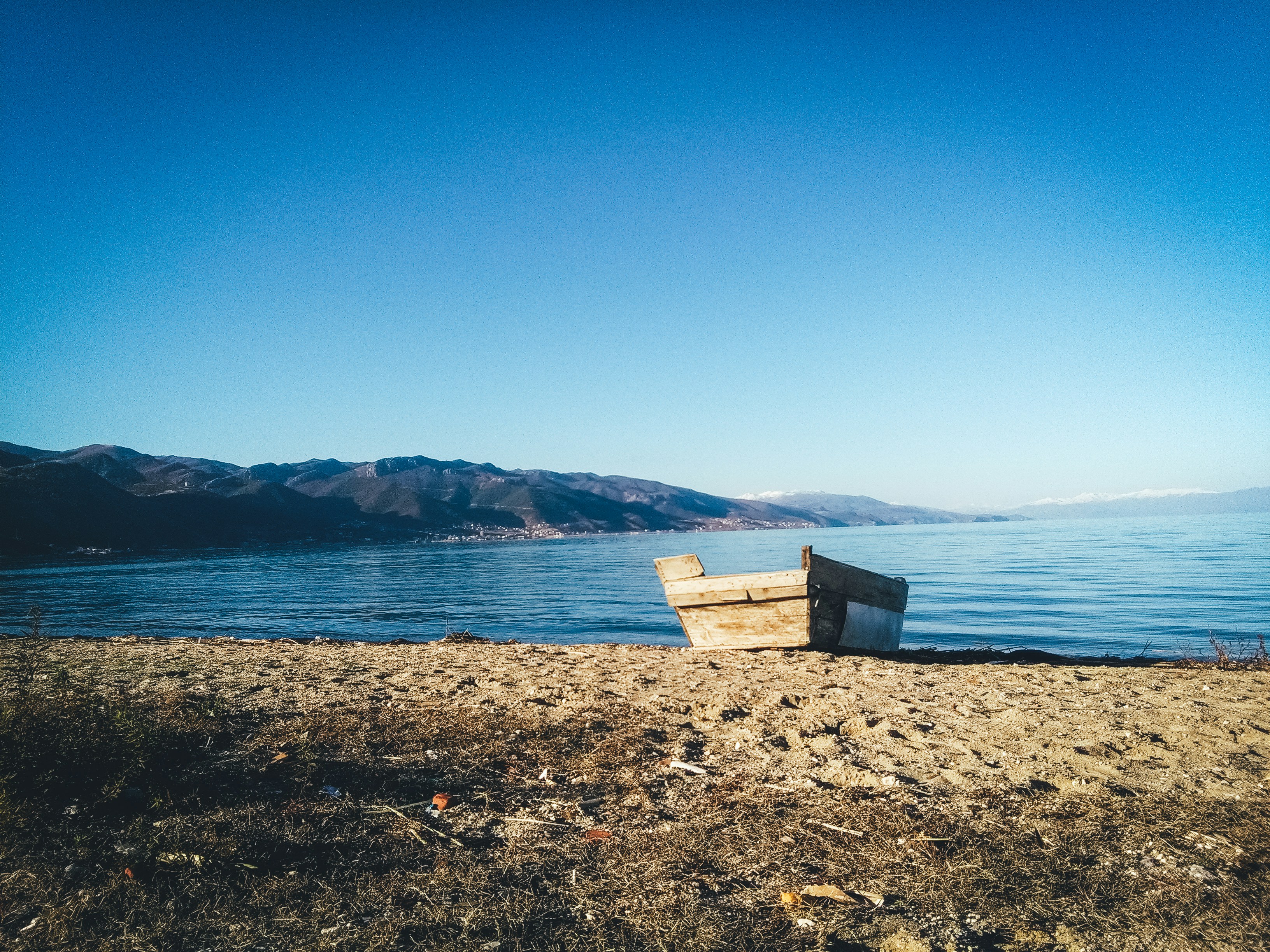 Two weathered boats rest on a rocky beach beside a calm bay, with distant mountains under a clear blue sky.