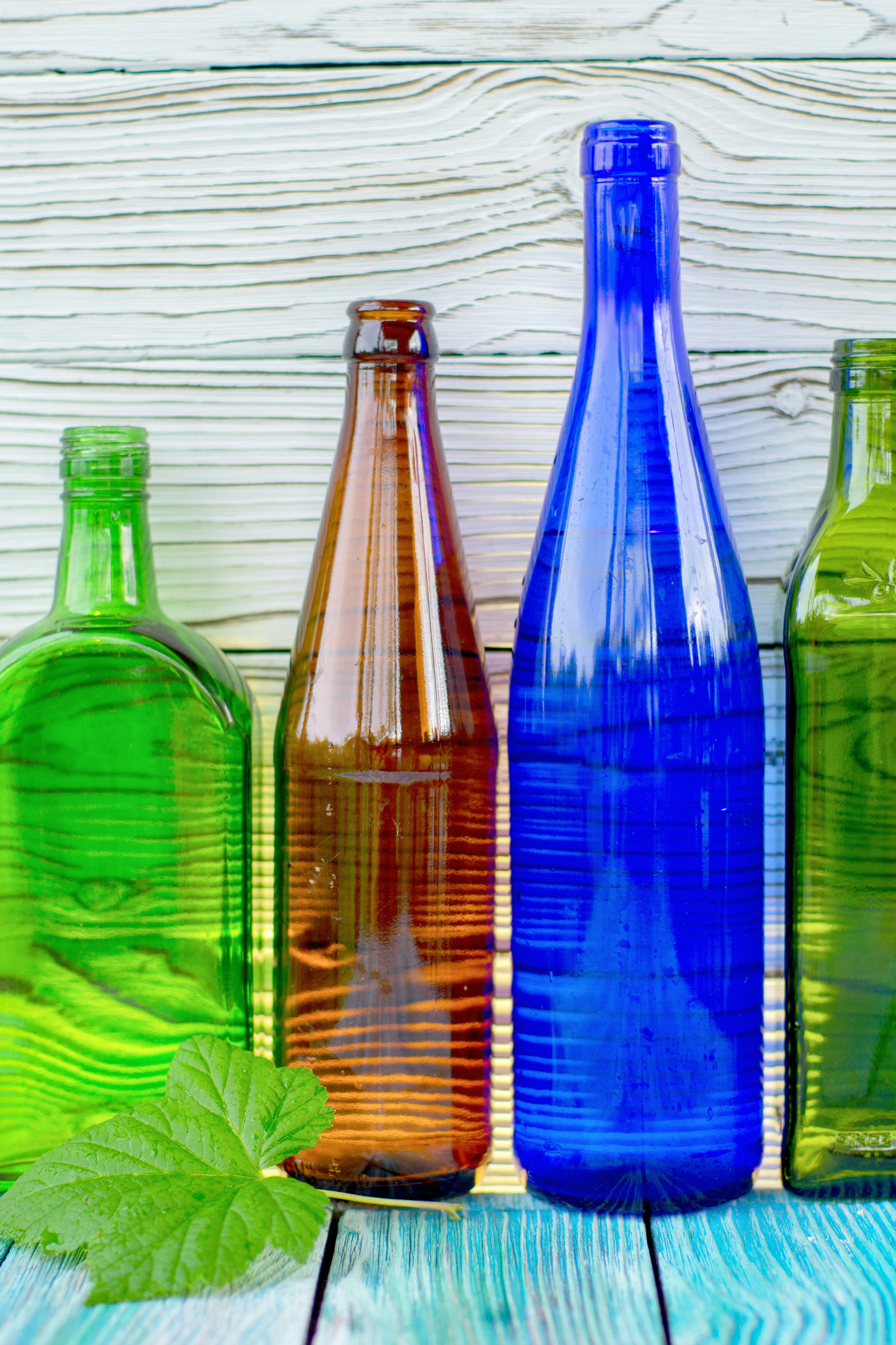 A lineup of colorful glass bottles in green, amber, blue, and clear, arranged against a textured wooden backdrop, accompanied by fresh green leaves.