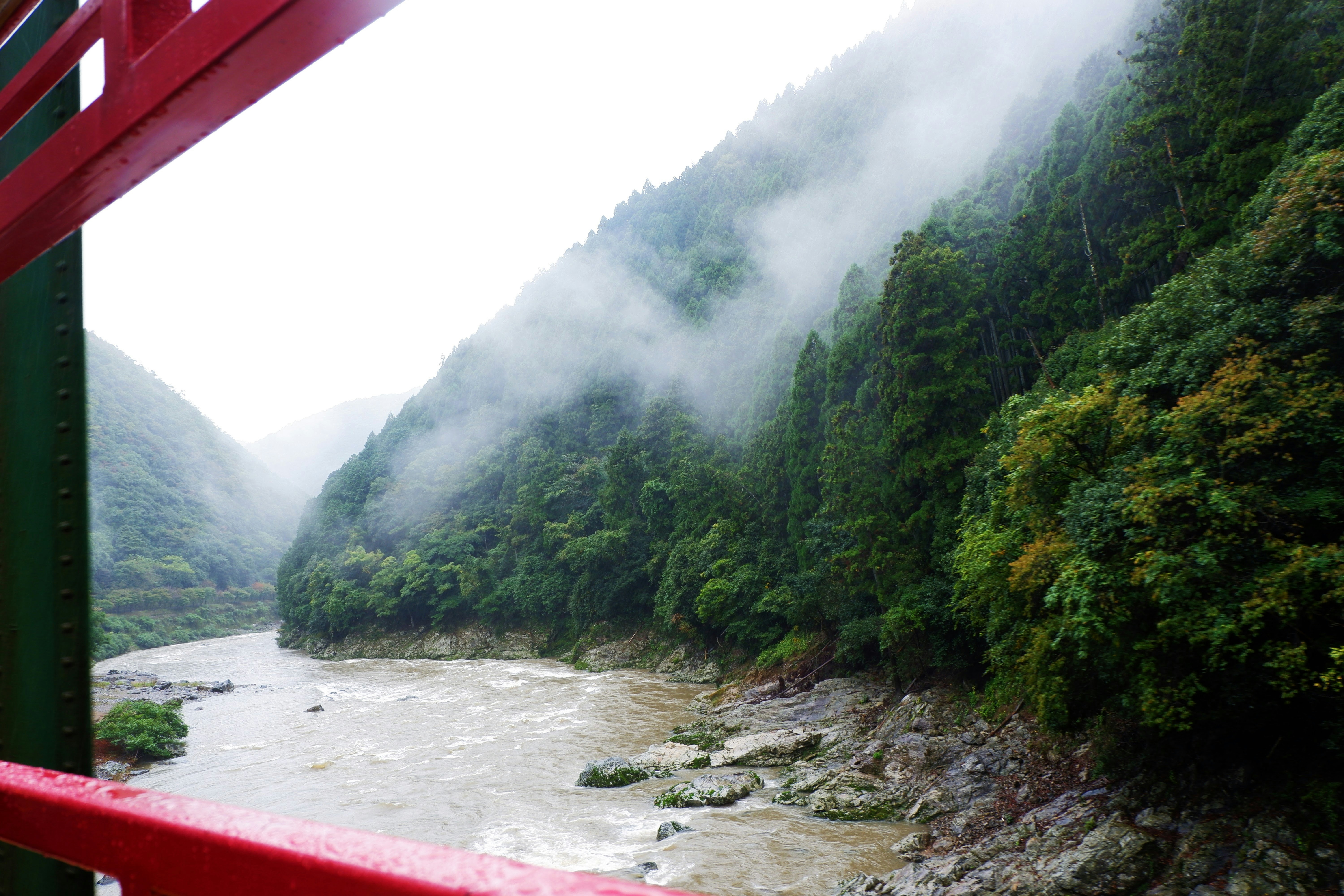A serene river meanders through lush green mountains, shrouded in mist, framed by a vibrant red bridge. The scene captures the tranquil essence of nature's beauty.