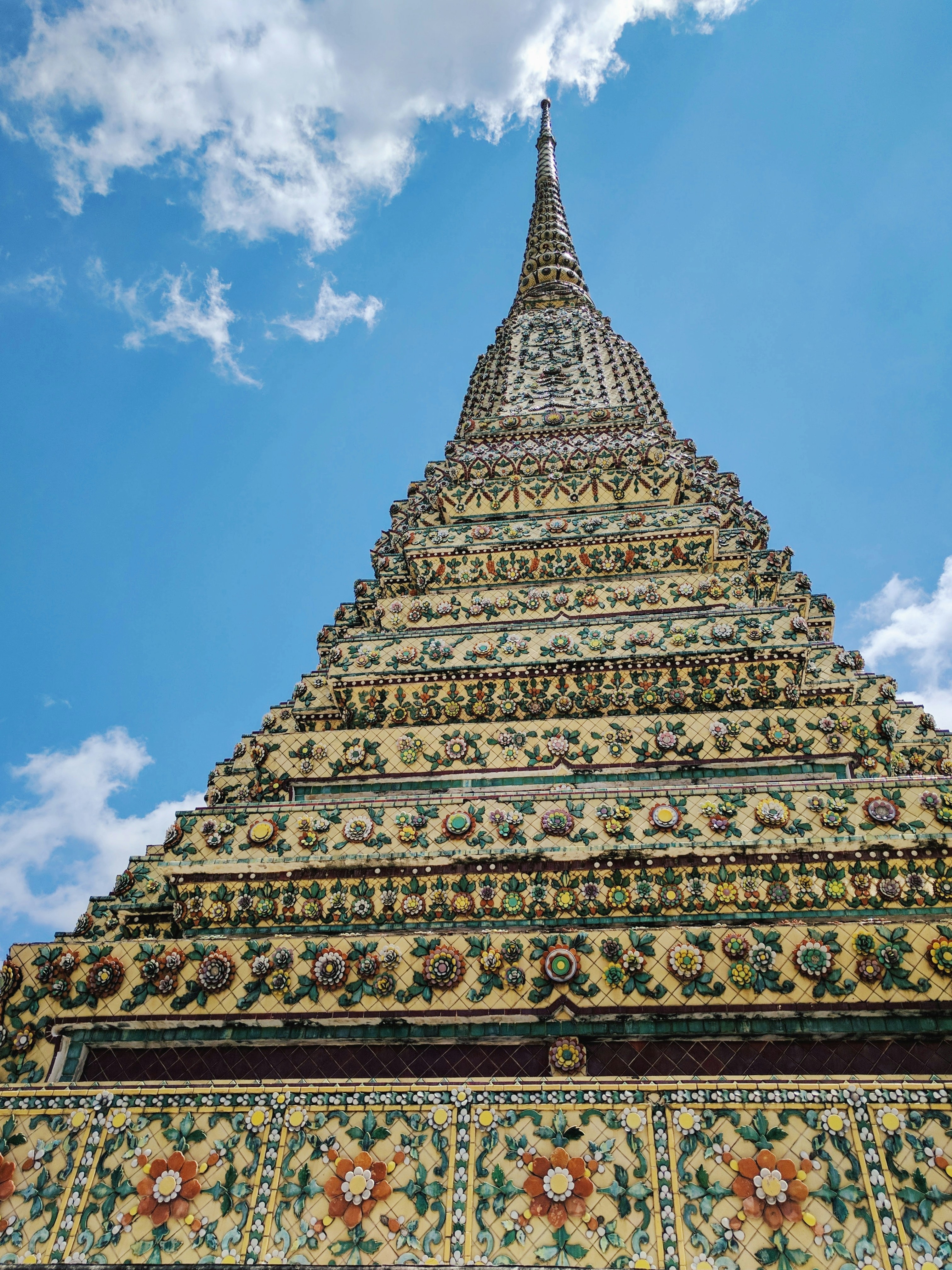 Ornate mosaic tiles cover a towering Thai temple stupa, rising toward a bright blue sky in a daytime photograph.