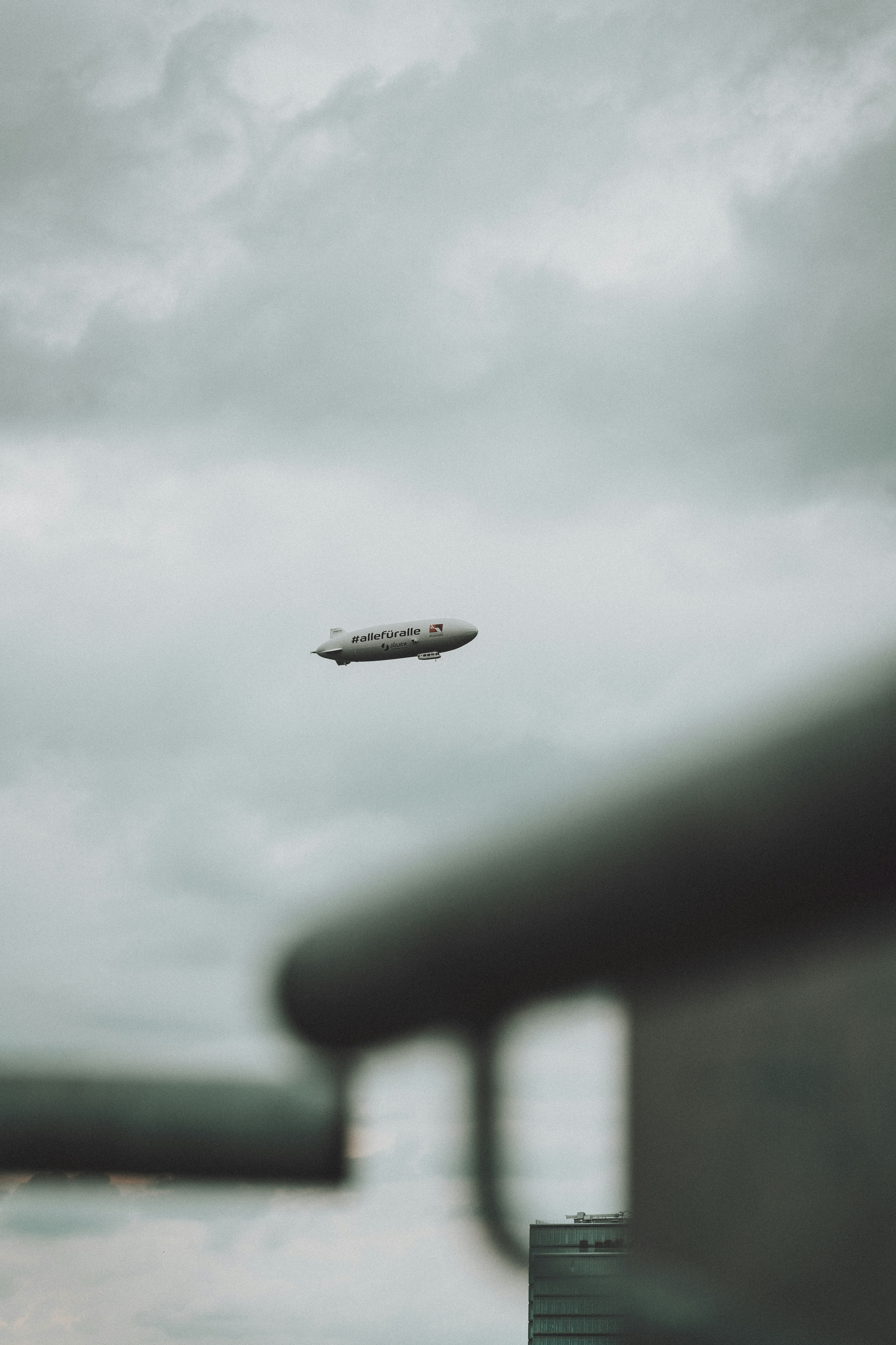 A zeppelin glides through overcast skies, partially obscured by a railing in the foreground. The scene captures a moment of tranquility amidst urban life.