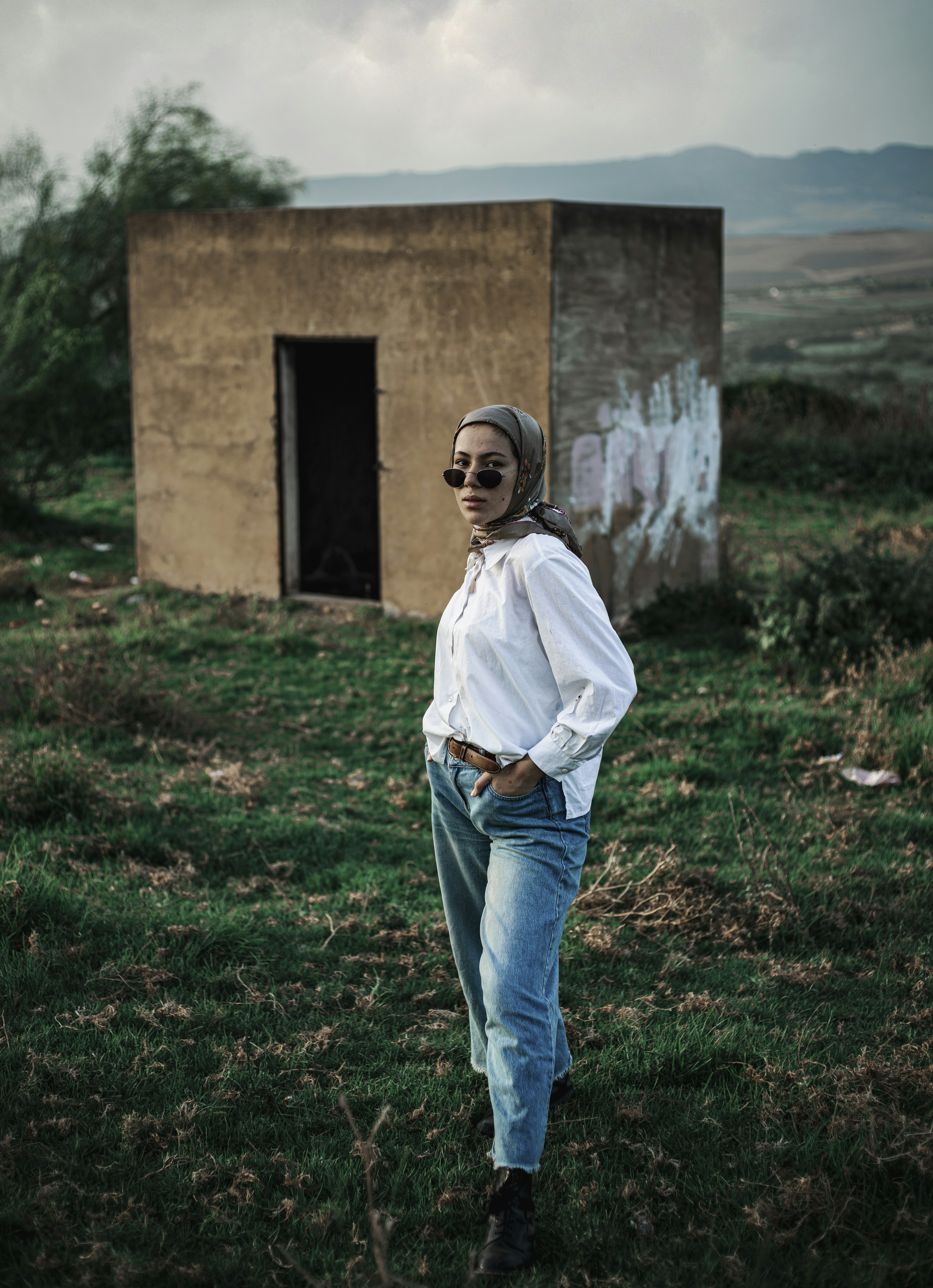 Fashionable individual in a white shirt and sunglasses poses confidently in front of a weathered structure in a lush green landscape.