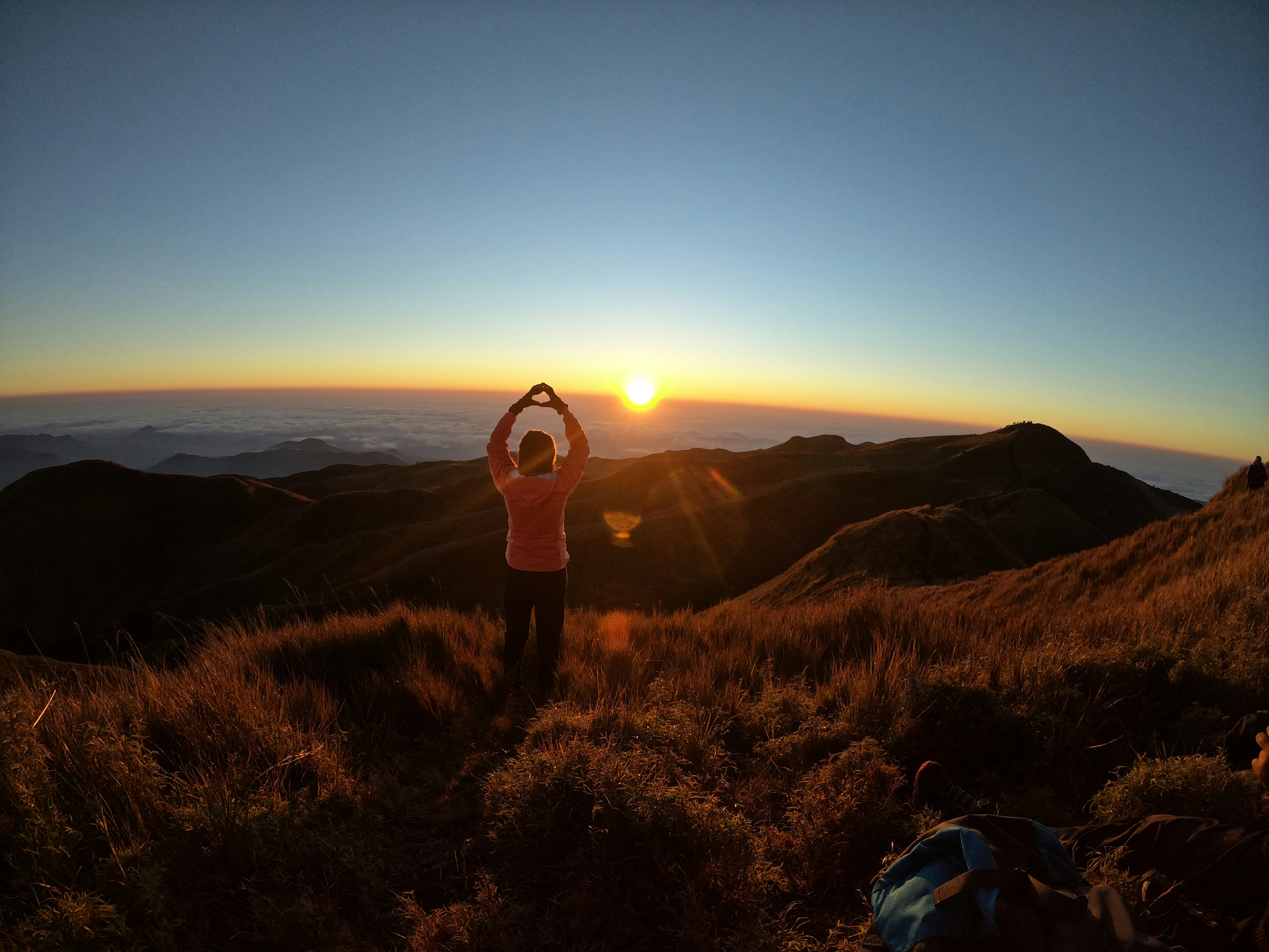 Sunset over a rugged ridge with a hiker in a pink jacket raising arms overhead to encircle the setting sun.