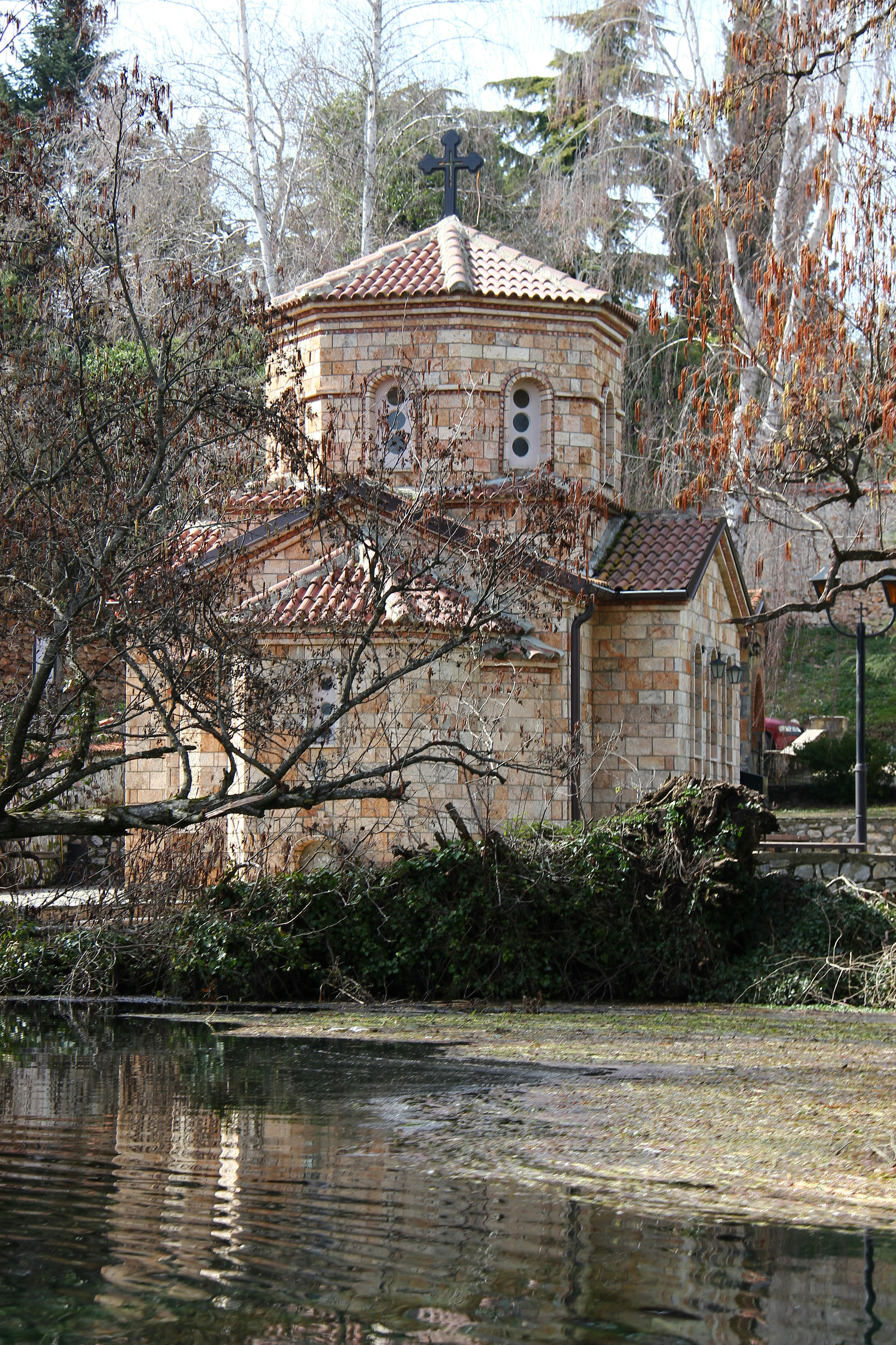 Charming stone chapel nestled among bare trees, reflecting in the tranquil waters nearby.