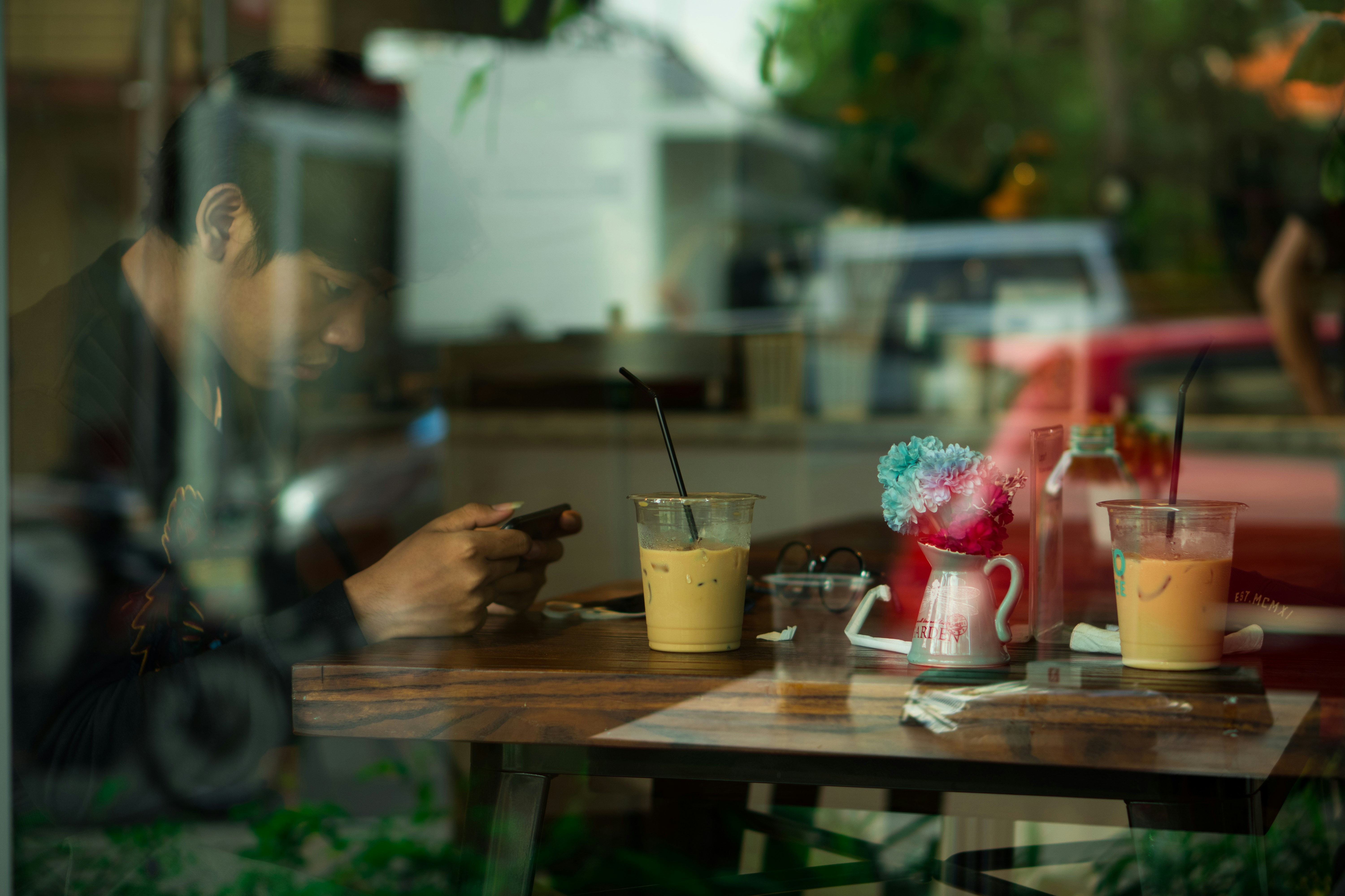 Man seated at a café table with drinks and a phone, seen through a reflective glass window.