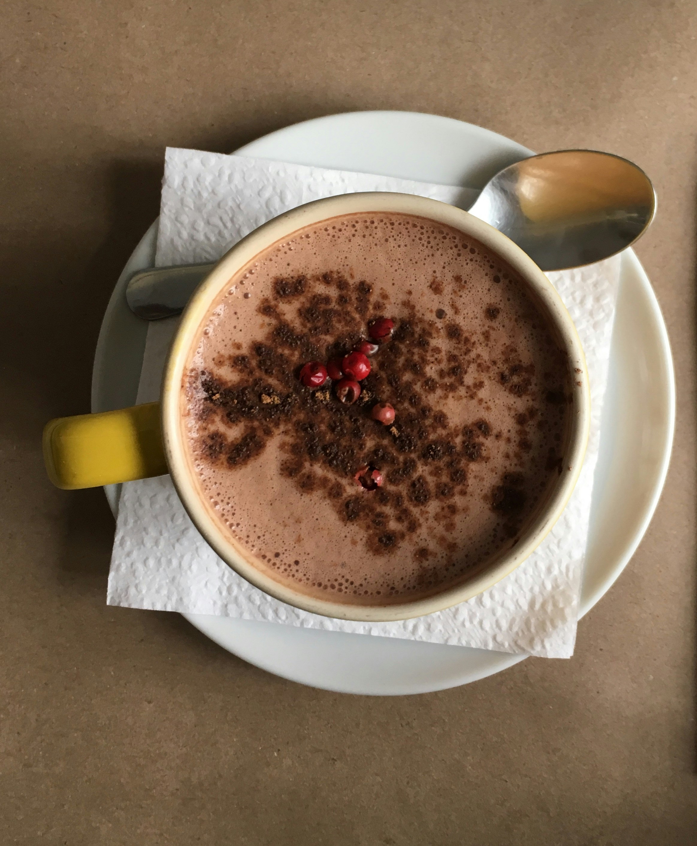 Rich cocoa topped with a sprinkle of spices and pink peppercorns, served in a vibrant yellow cup on a white plate with a spoon.