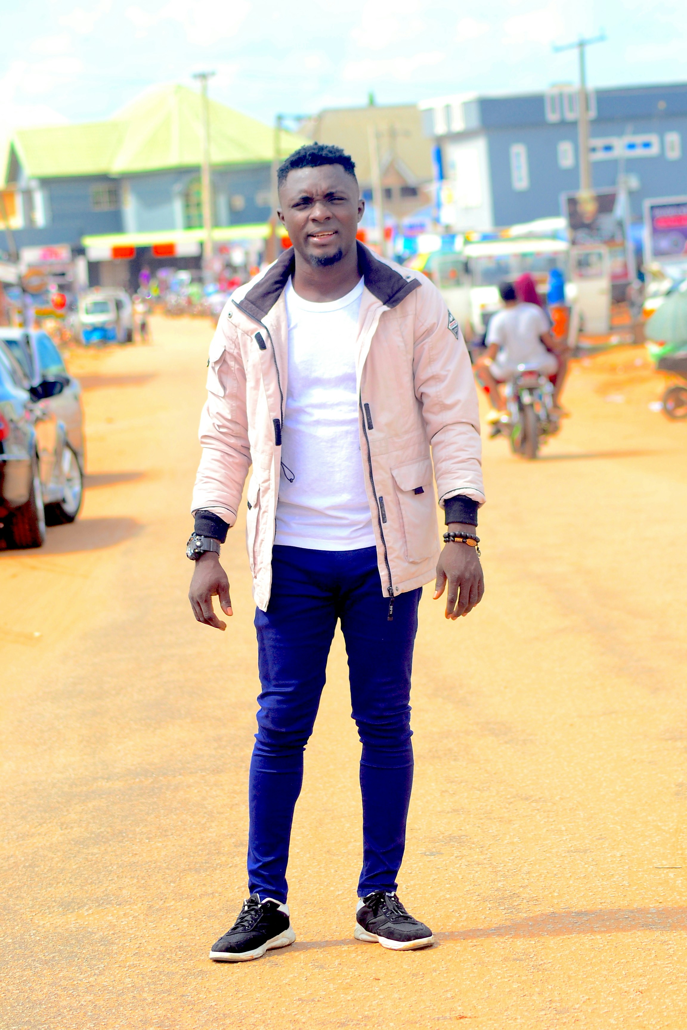 A young man stands confidently on a bustling street, dressed casually in a light jacket and sneakers, surrounded by the vibrant activity of city life.