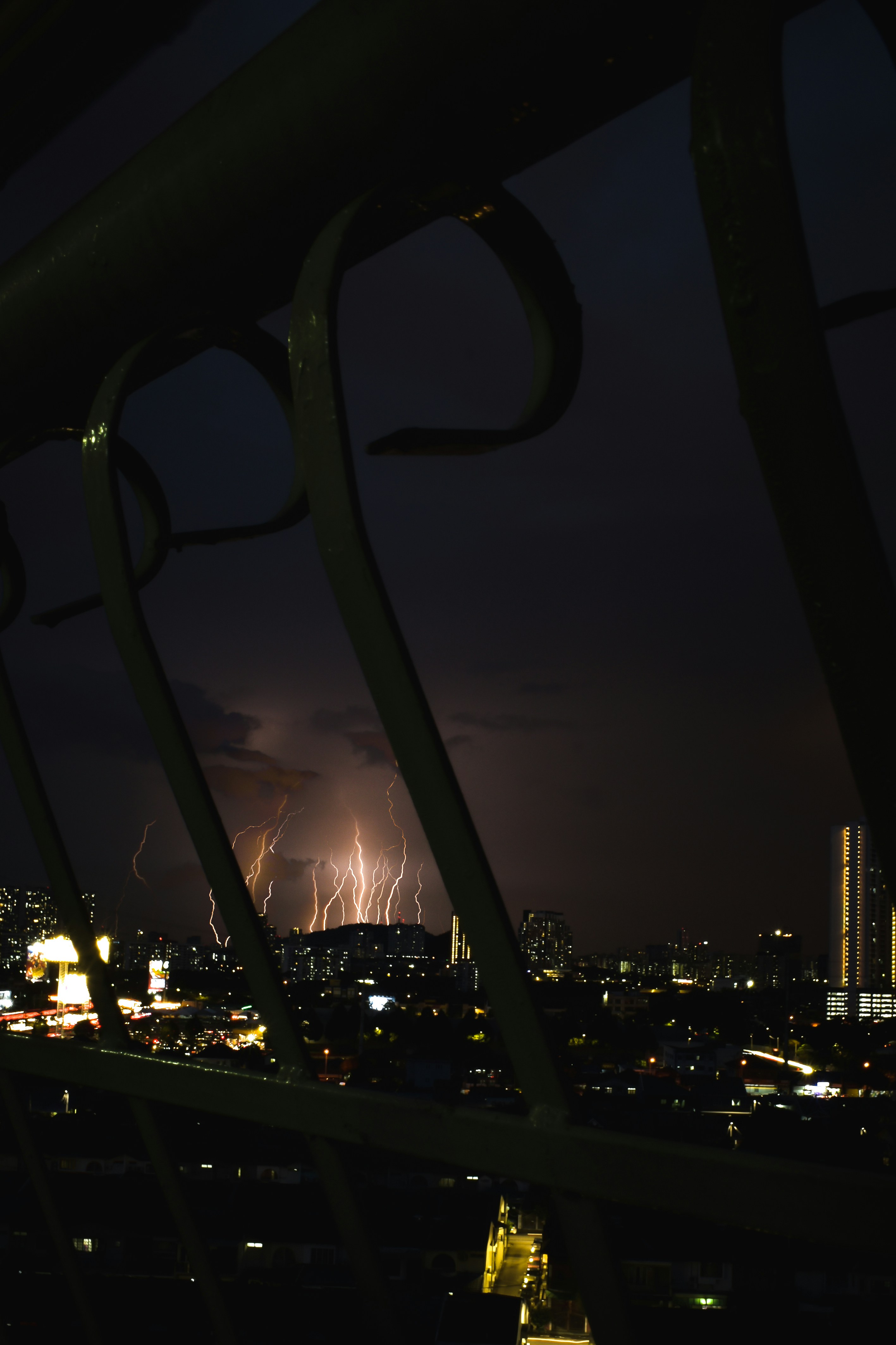 Lightning strikes illuminate the skyline, framed by a balcony railing during a thunderstorm. The vibrant city lights contrast with the dark clouds above.