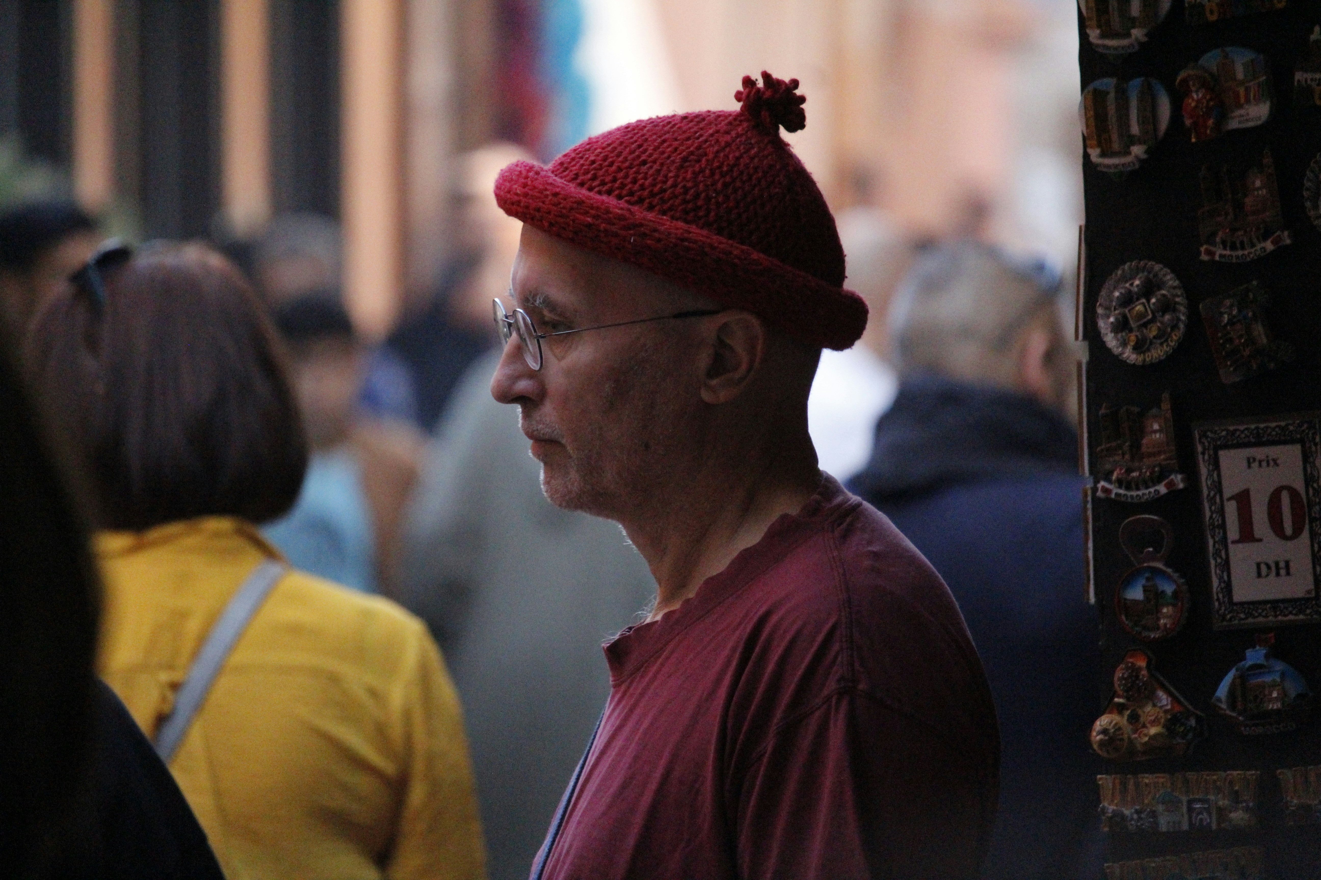 Profile of a man wearing a red knitted hat, standing amidst a bustling market scene. The background is filled with blurred figures, emphasizing his solitude.