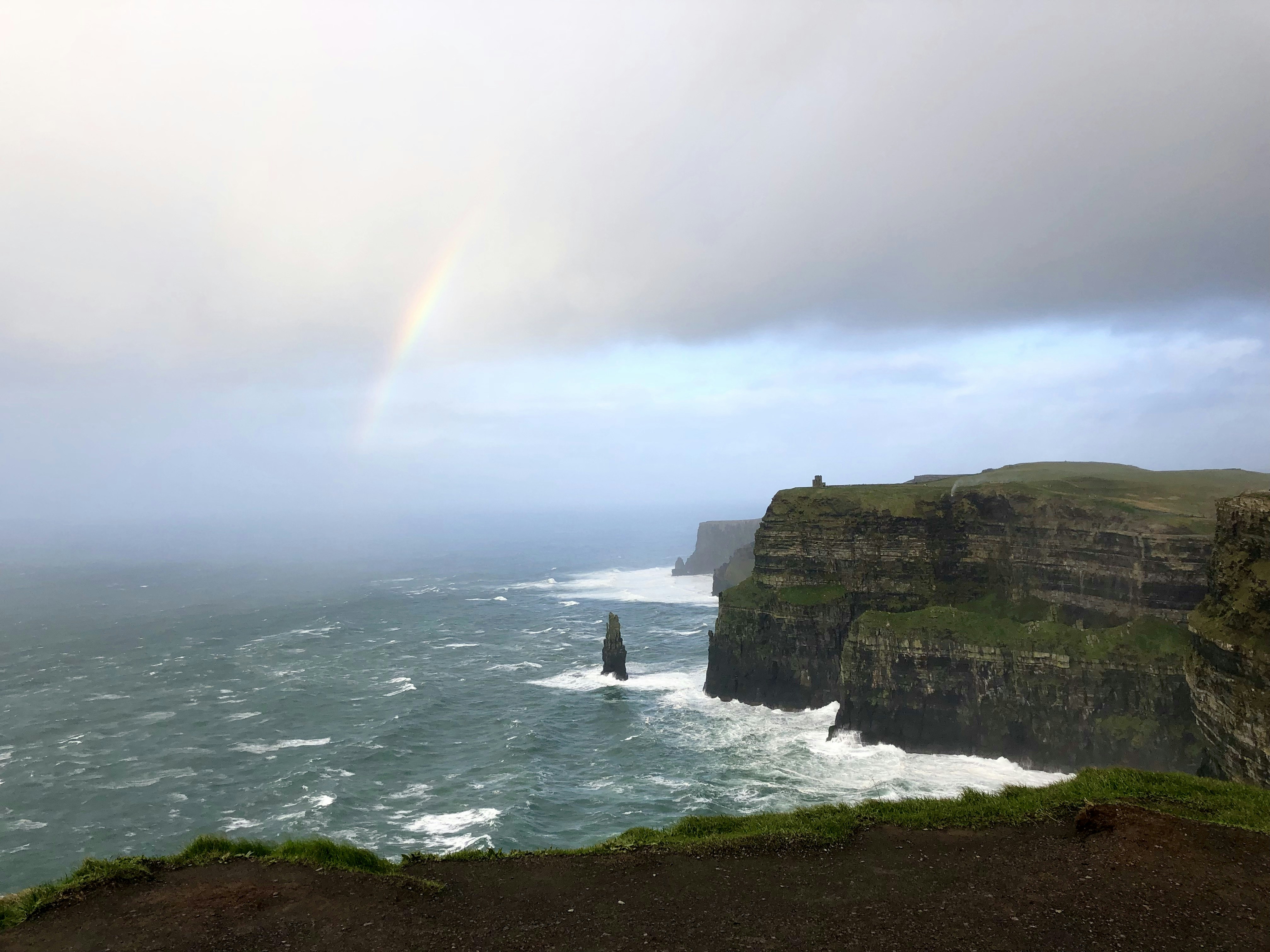 Cliffs meet the turbulent sea under a cloudy sky with a faint rainbow arching above.