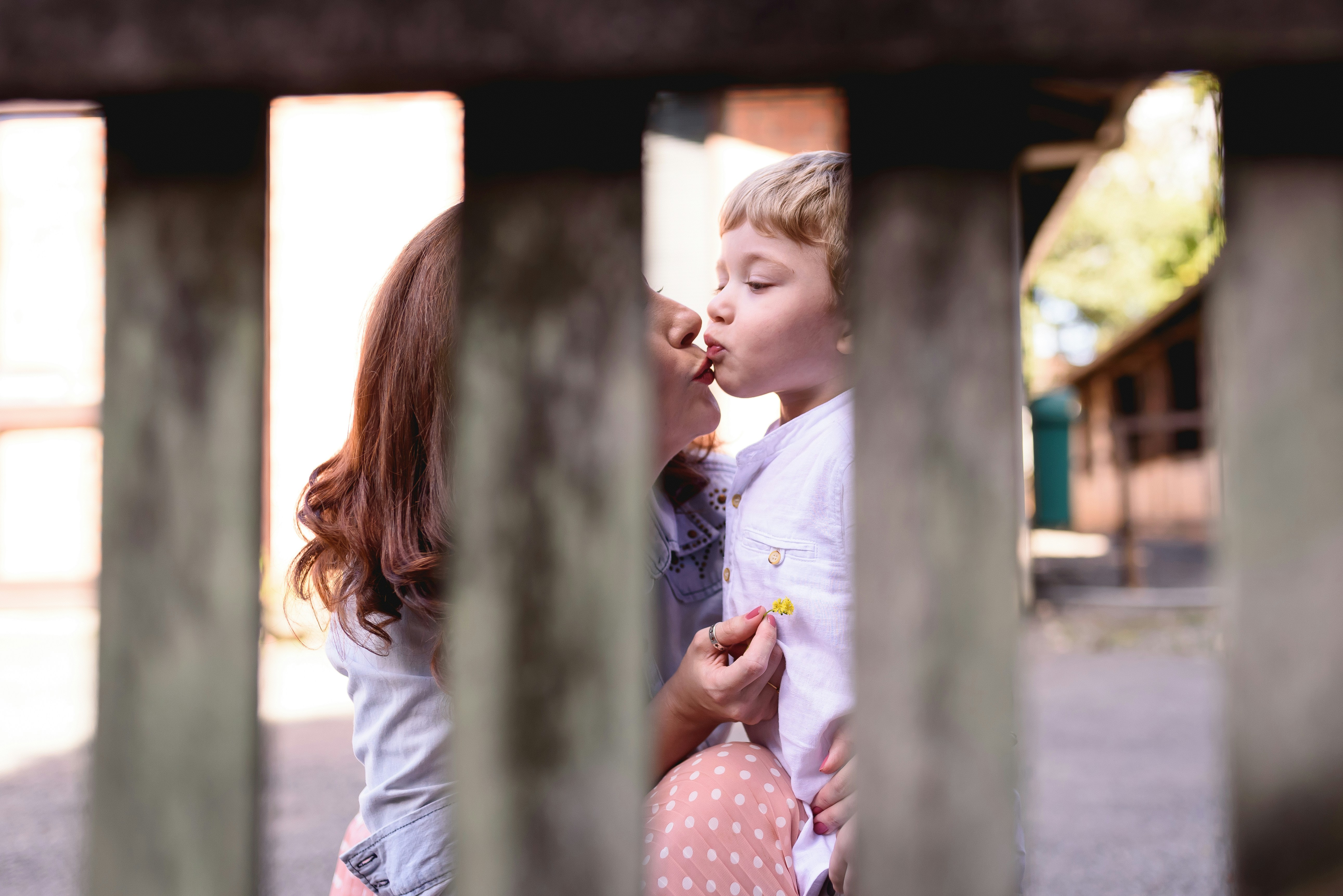 Child in a white shirt holding and kissing a baby in a pink and white polka dot outfit, viewed through wooden slats.