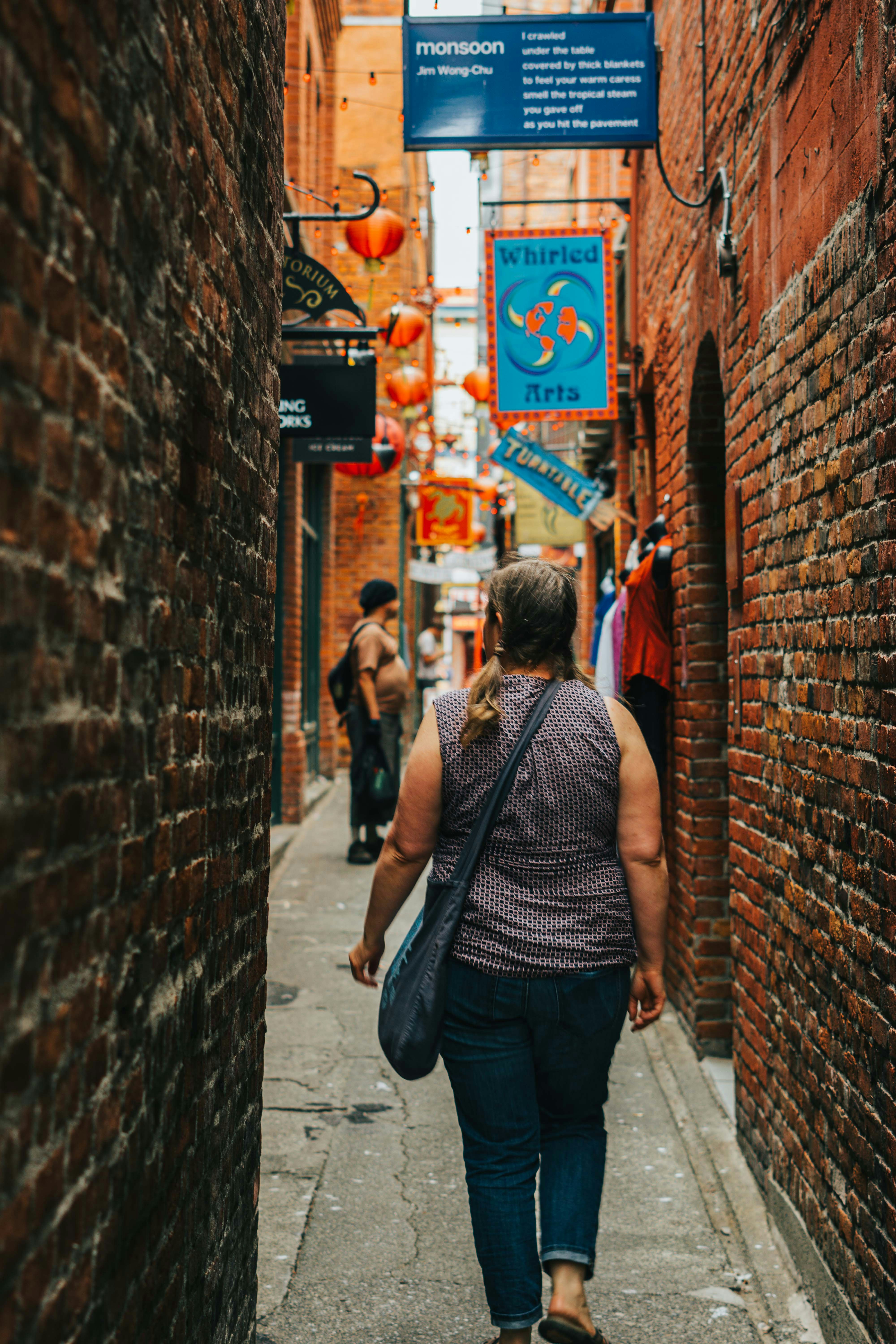 A woman strolls through a narrow alley adorned with colorful signs and lanterns, showcasing the lively atmosphere of a bustling marketplace.