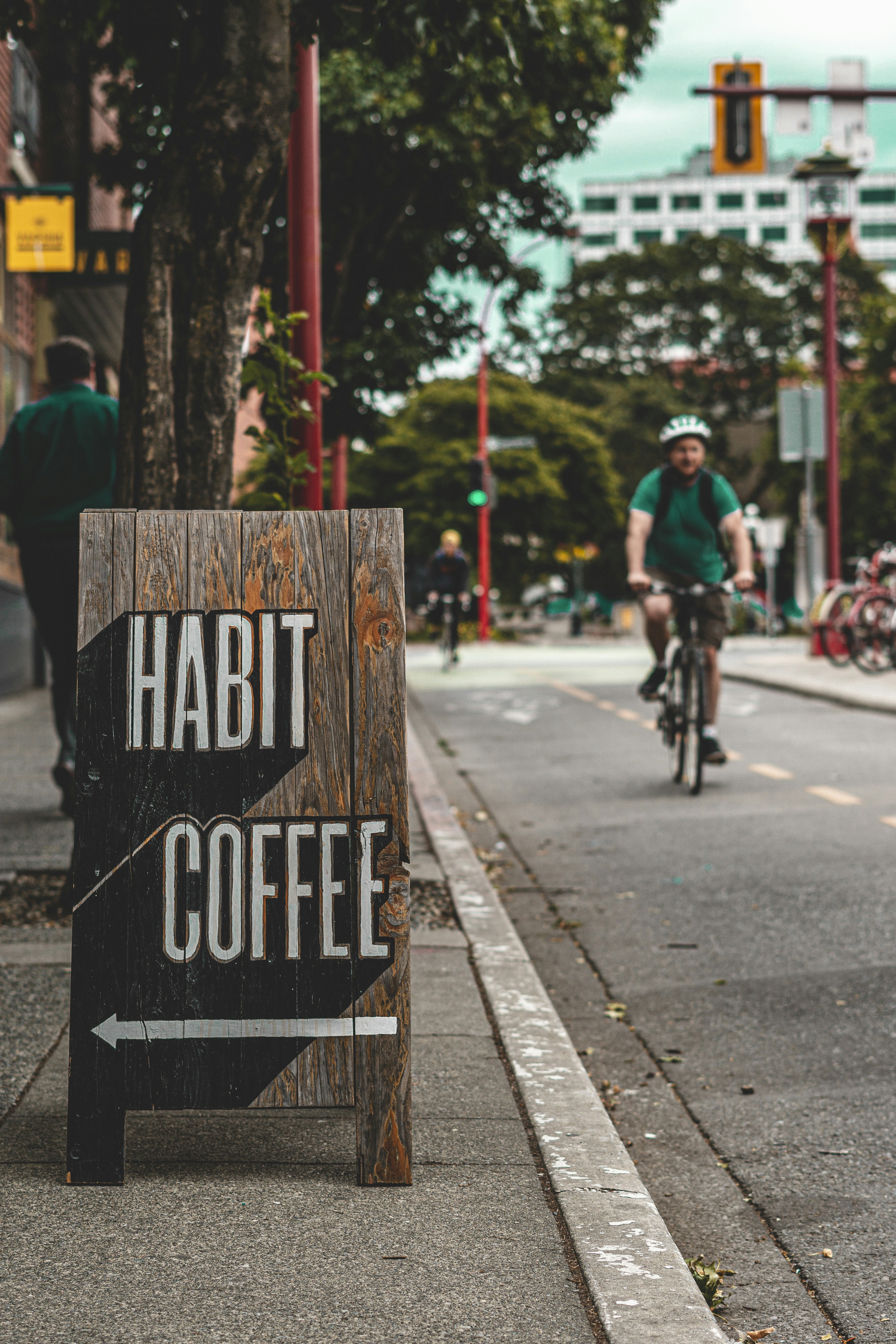 Wooden sign for 'Habit Coffee' prominently displayed on a city sidewalk, with a cyclist passing by. The scene captures the essence of urban life.
