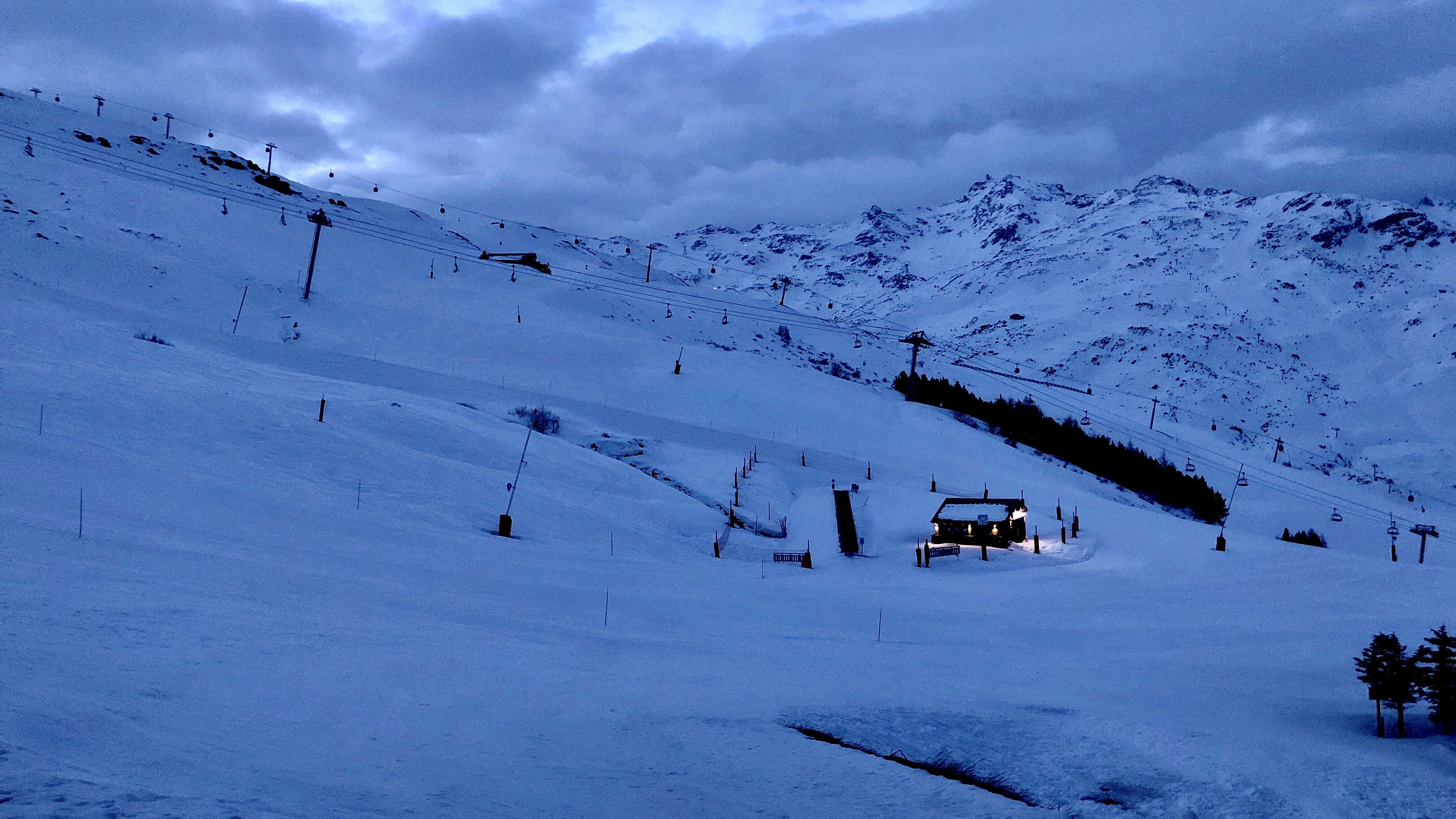 A lone lodge illuminated against the backdrop of a vast, snowy landscape under a twilight sky, with ski lifts standing silently nearby.