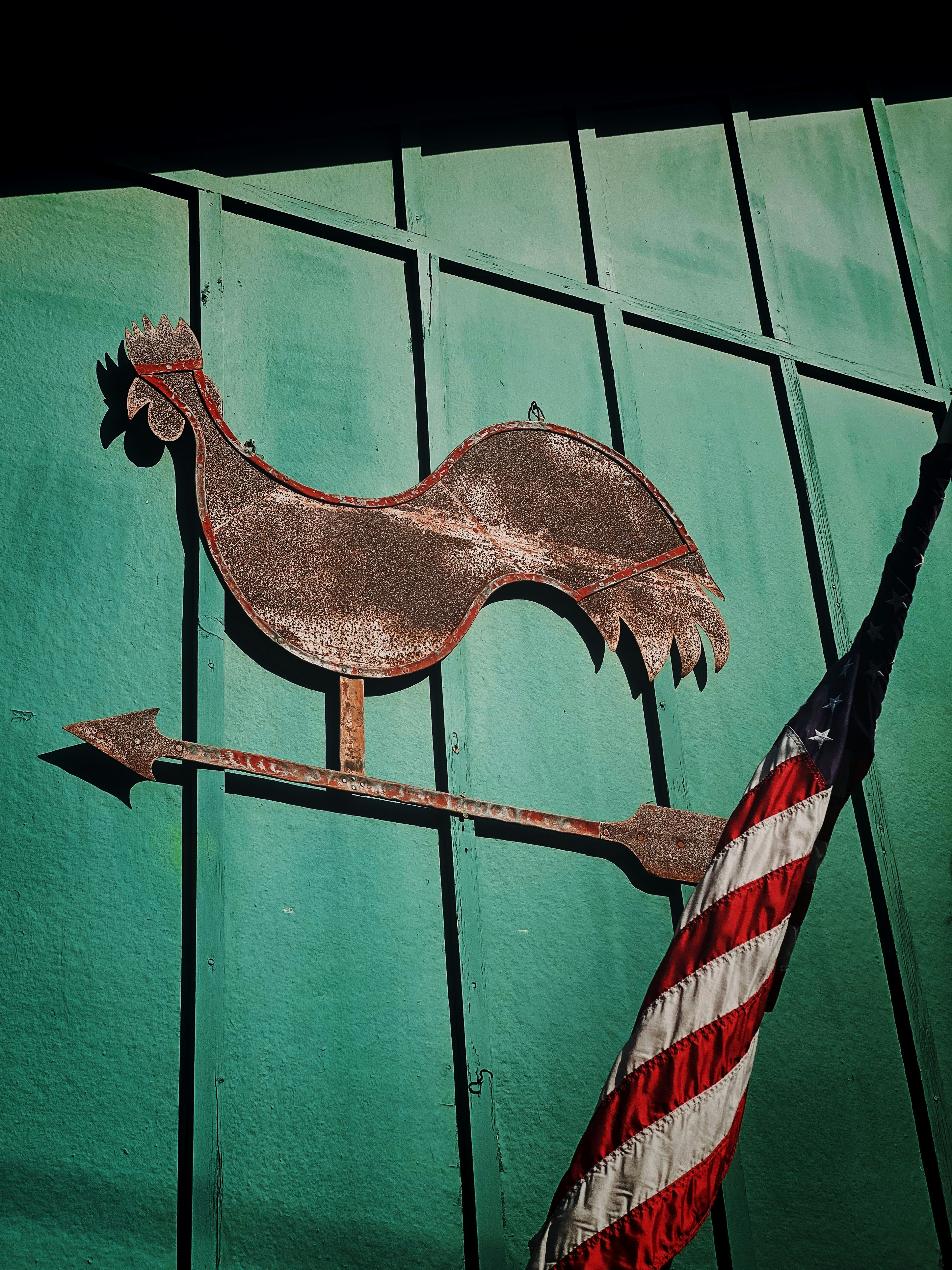 Weather vane shaped like a rooster mounted on a vibrant green wall, accompanied by a tattered American flag.