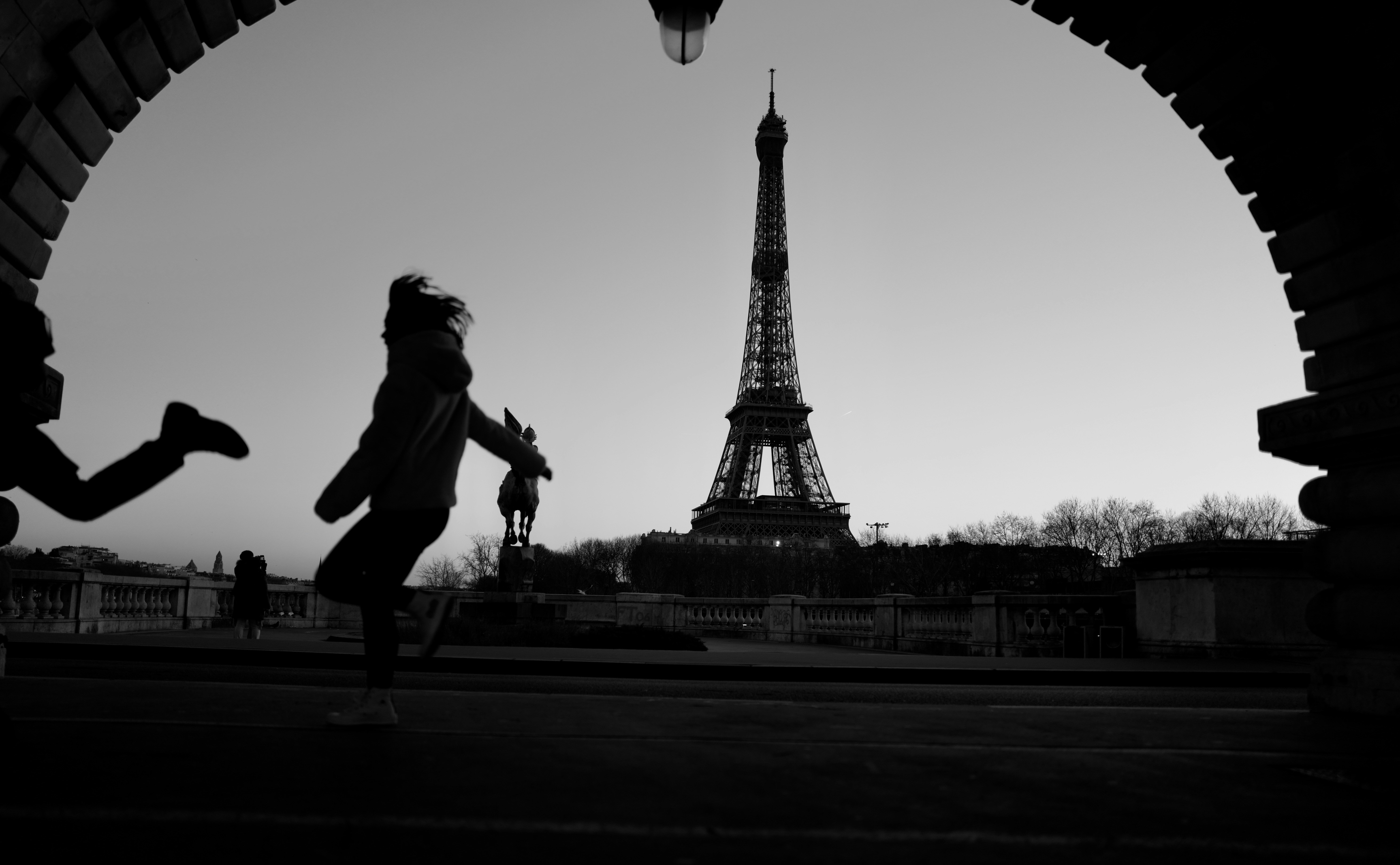 Two figures playfully dash through an archway, silhouetted against the iconic Eiffel Tower at twilight.