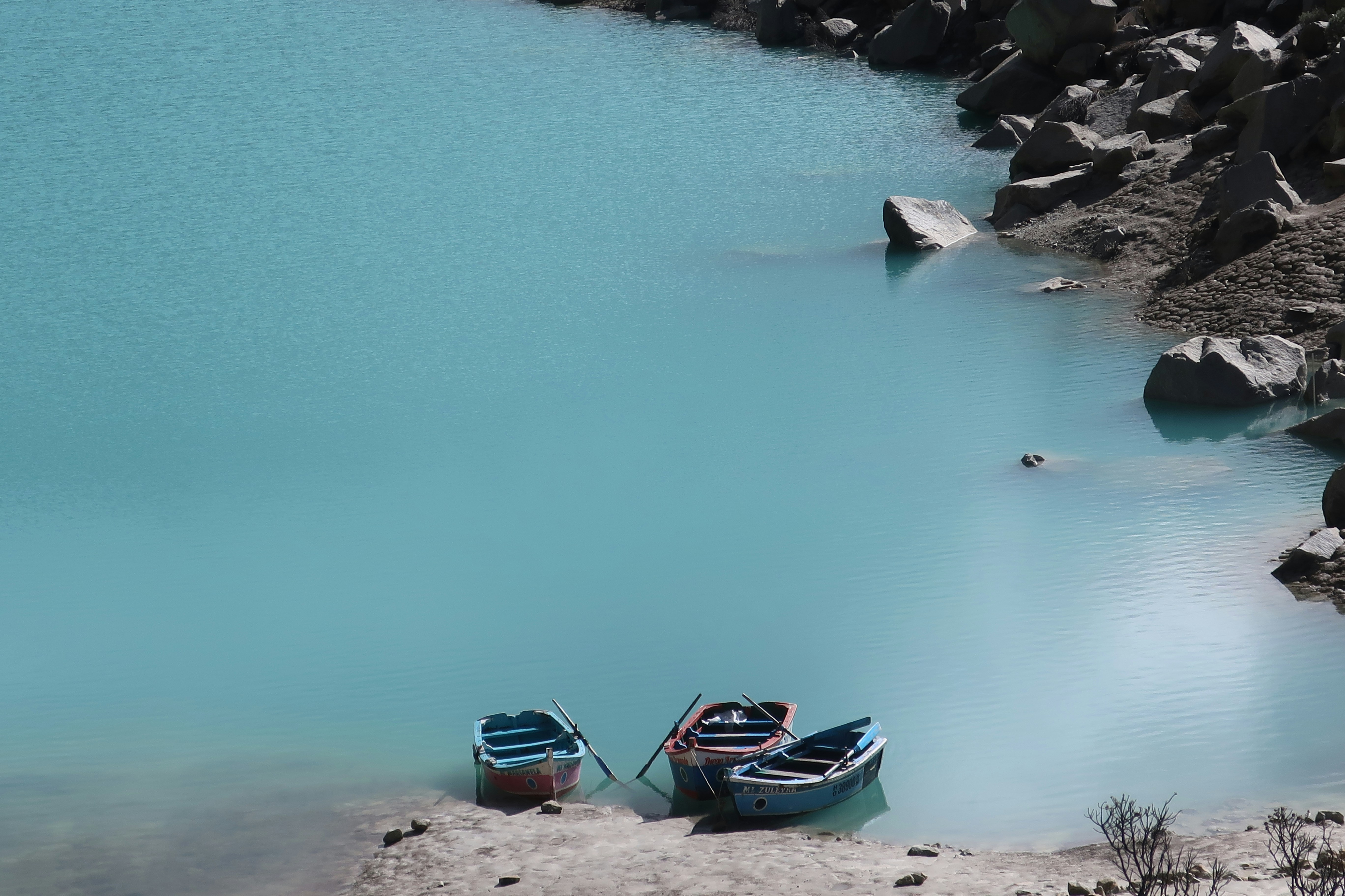 Three small boats anchored on the edge of a tranquil, turquoise lake surrounded by rocky terrain.
