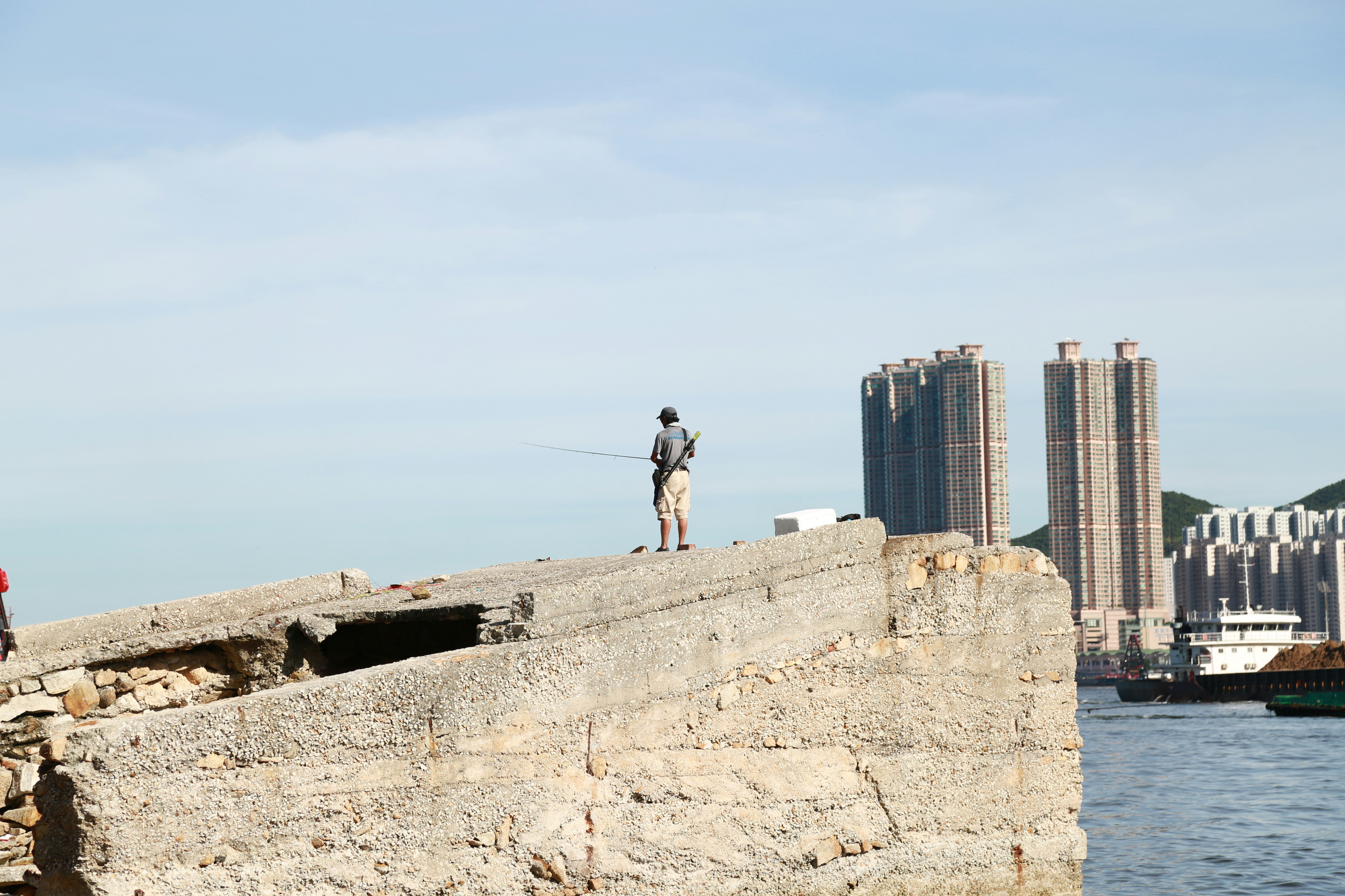Edge of Tides: A Fisherman on Weathered Concrete with a City Skyline Backdrop