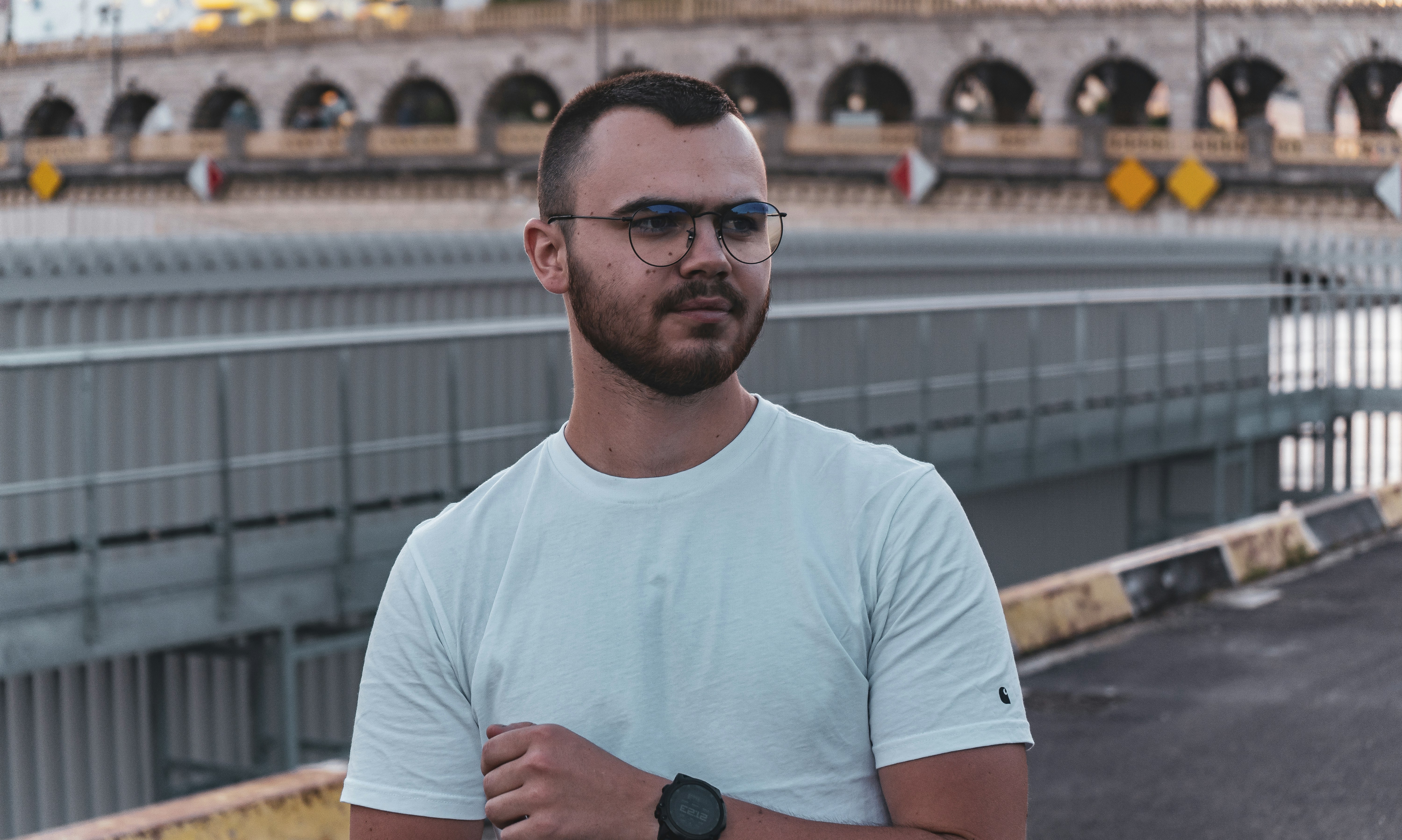Man wearing a white t-shirt and sunglasses standing in front of an urban overpass.