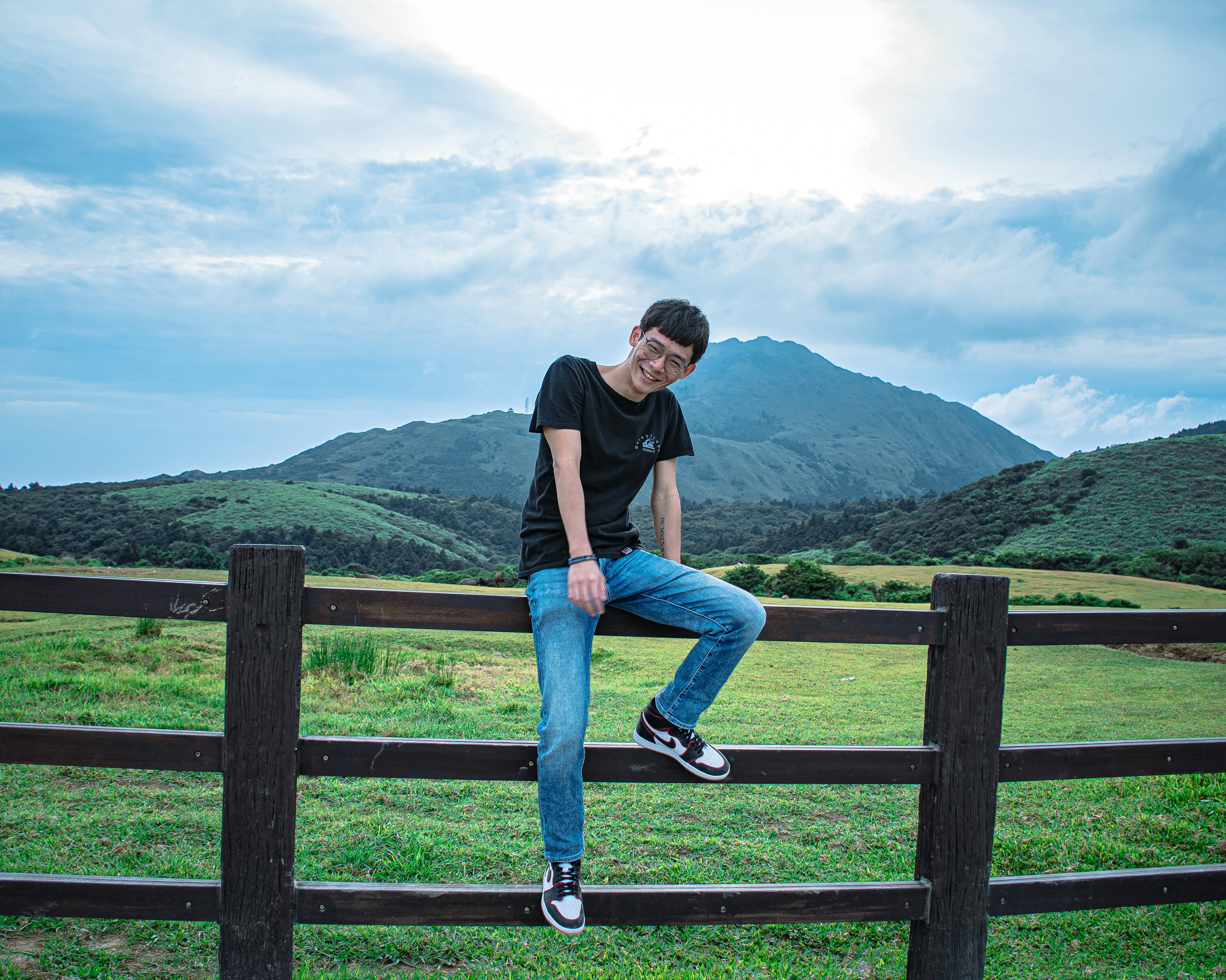 man in black shirt and blue denim jeans sitting on brown wooden fence during daytime