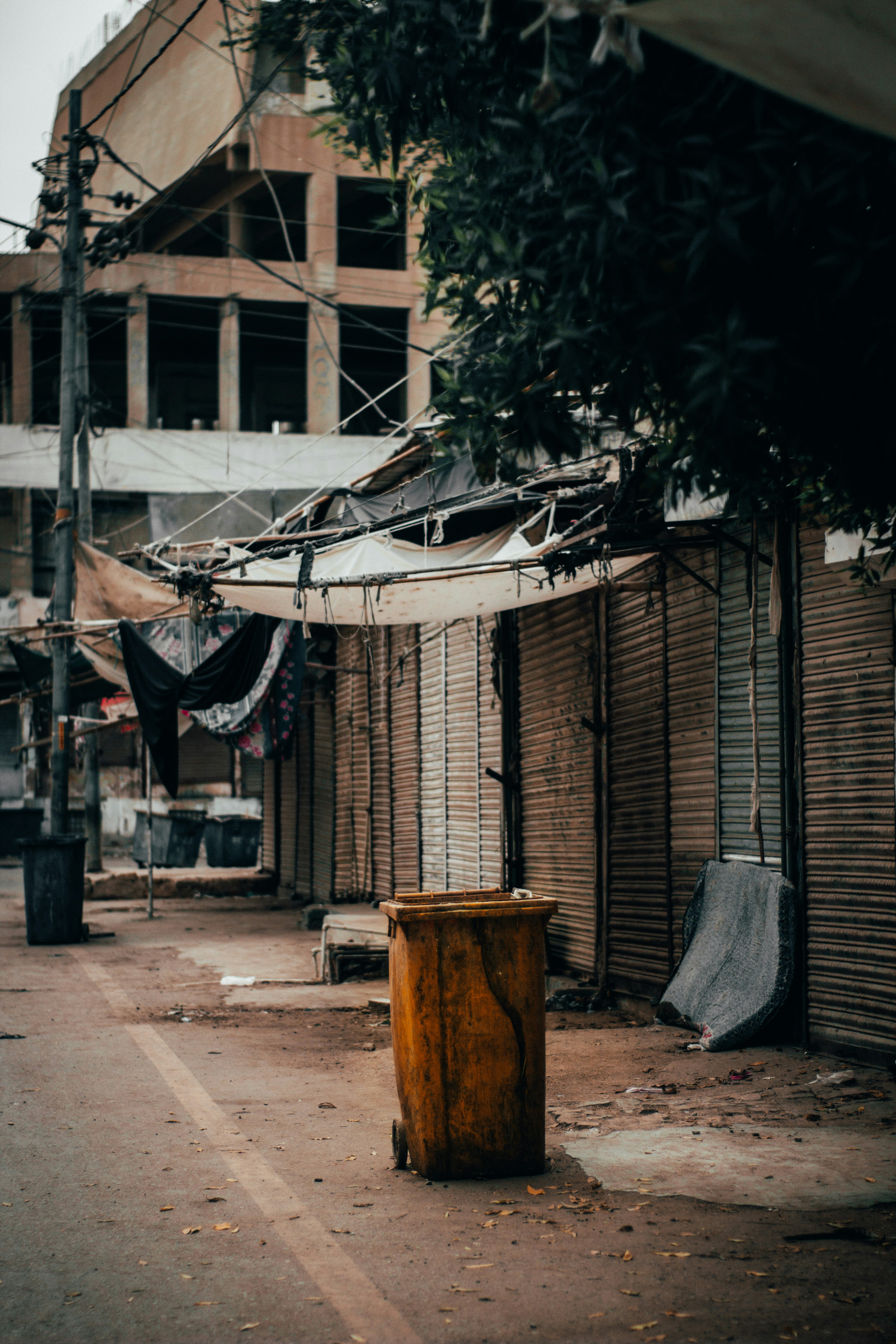 Brown wooden trash bin in a deserted street with closed market stalls and overhanging foliage.