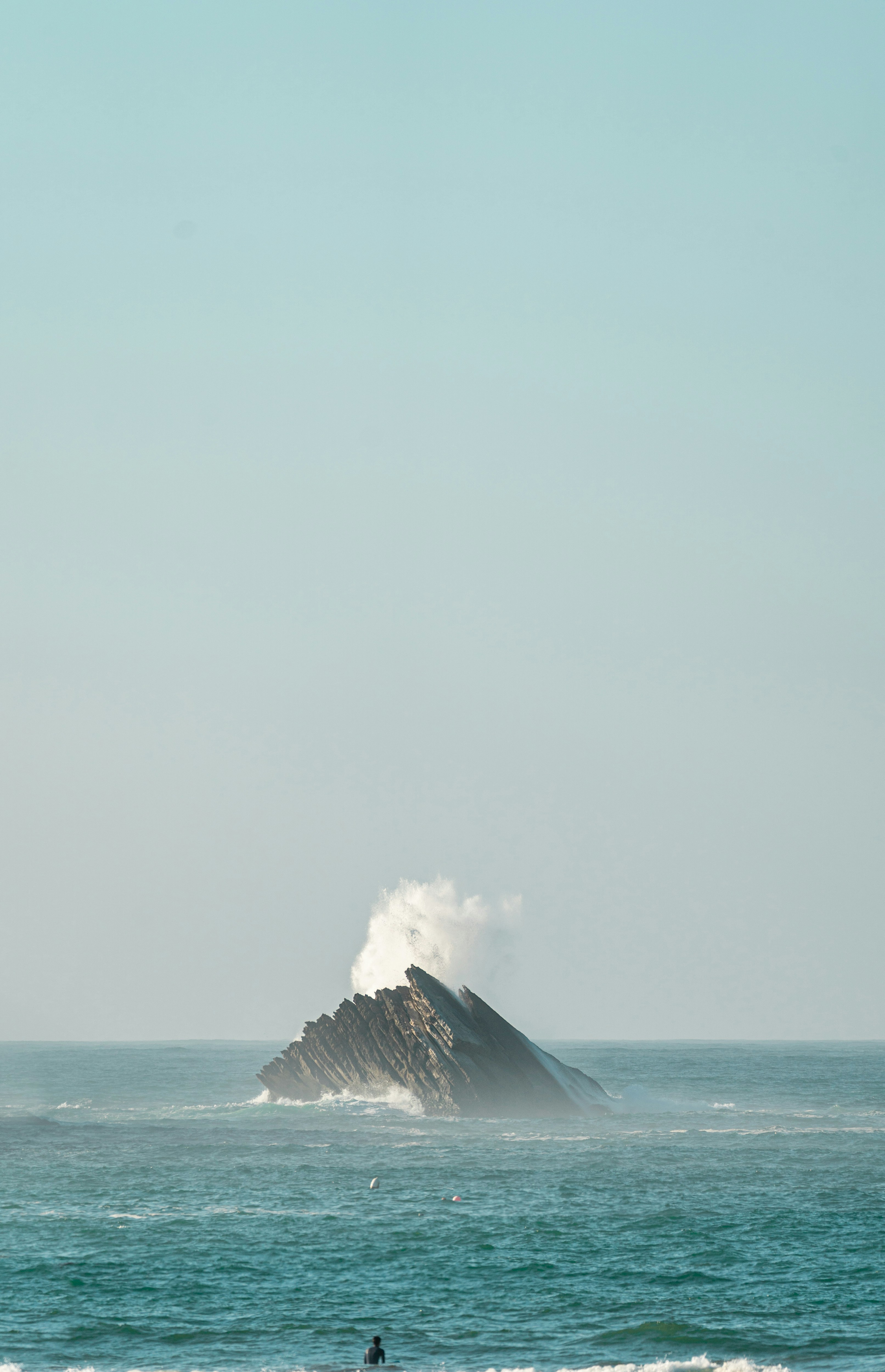A solitary rock formation rises from the calm sea, with a gentle plume of spray dancing in the air. The serene blue backdrop enhances the tranquil atmosphere.