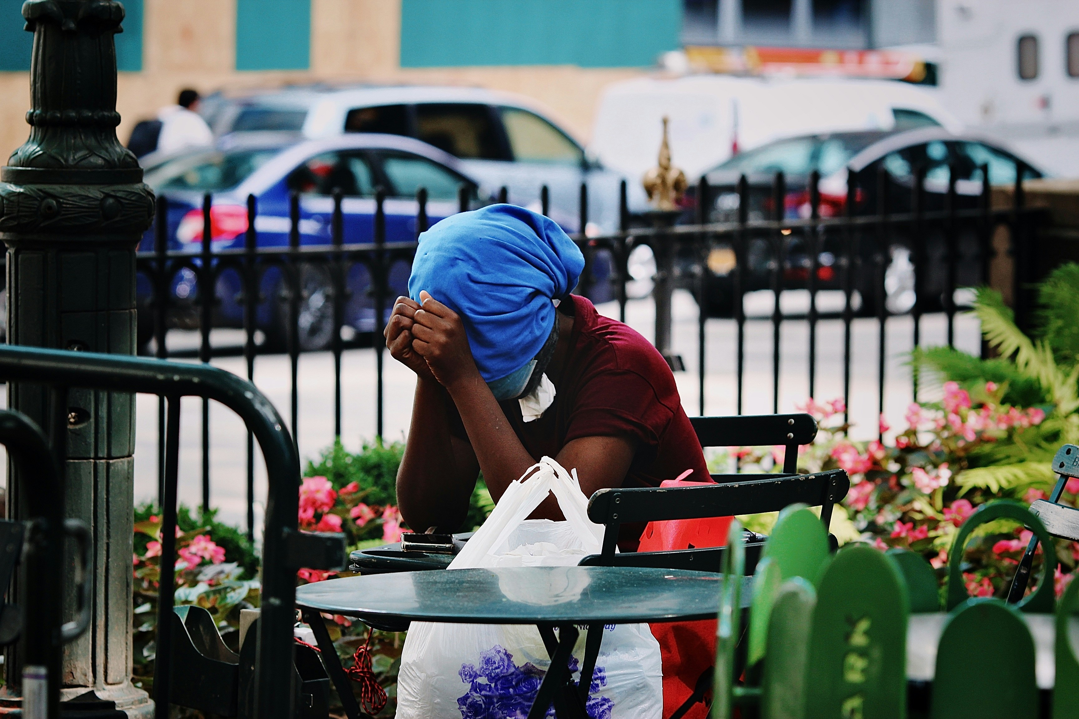 A person seated at a café table, head bowed and covered, surrounded by vibrant flowers and bustling city life.