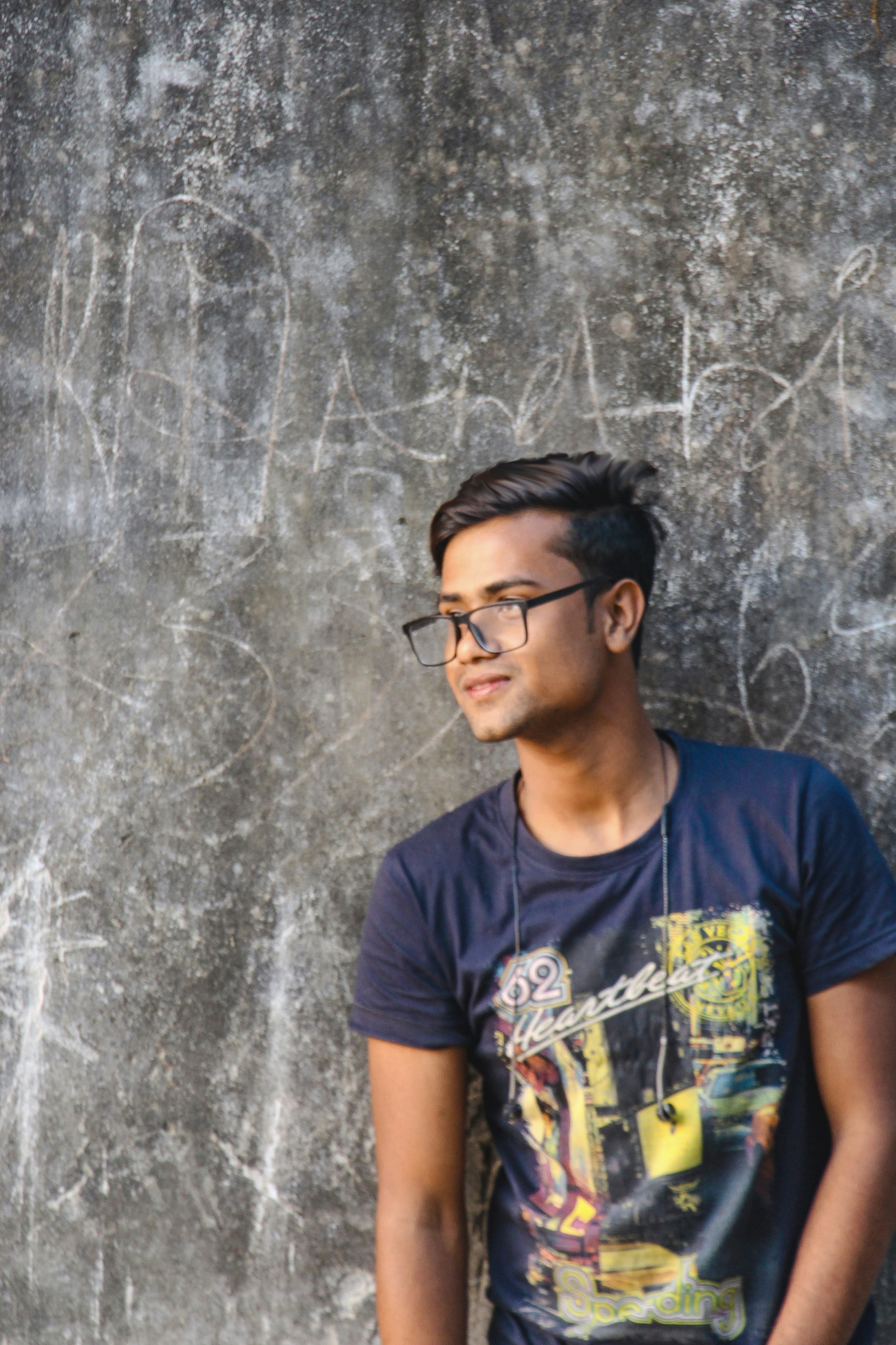 Young man in casual attire leans against a textured wall, exuding a relaxed demeanor. The subtle graffiti in the background adds an urban touch.