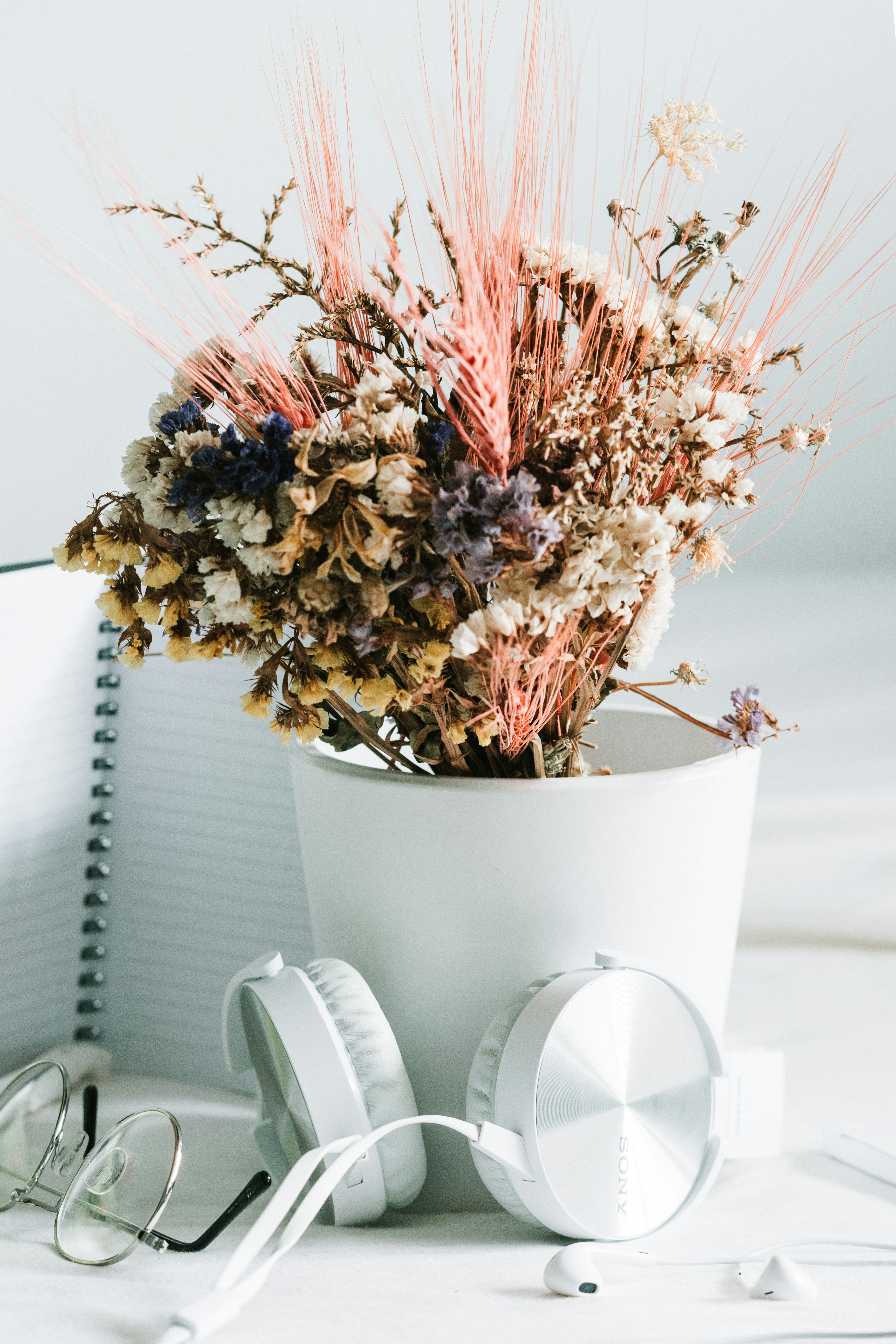 A delicate arrangement of dried flowers in a white pot, complemented by modern headphones and a notebook, creating a serene workspace aesthetic.
