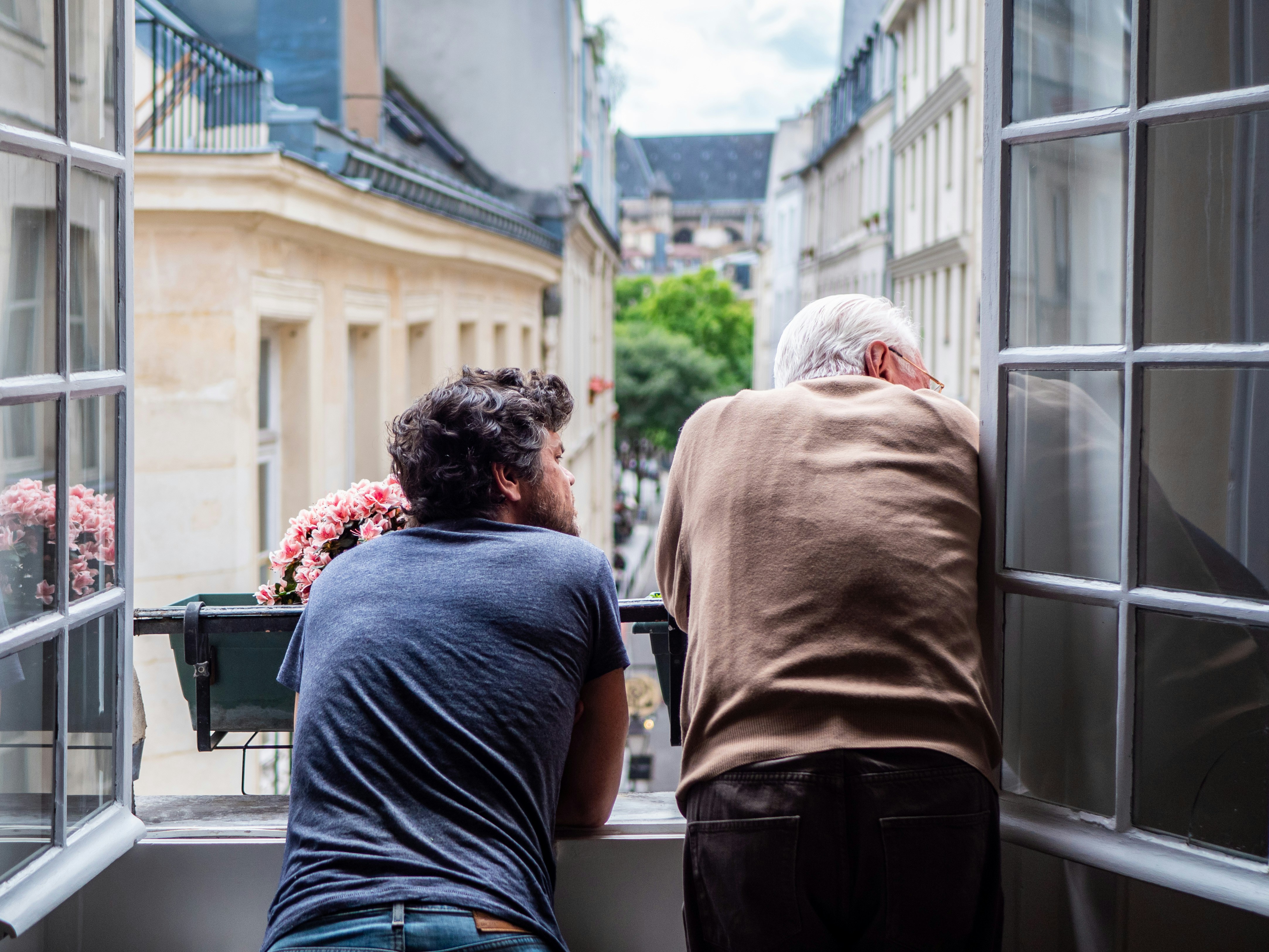 Two individuals leaning out of a window, observing the street below adorned with flowers. A glimpse of Parisian architecture frames the scene.