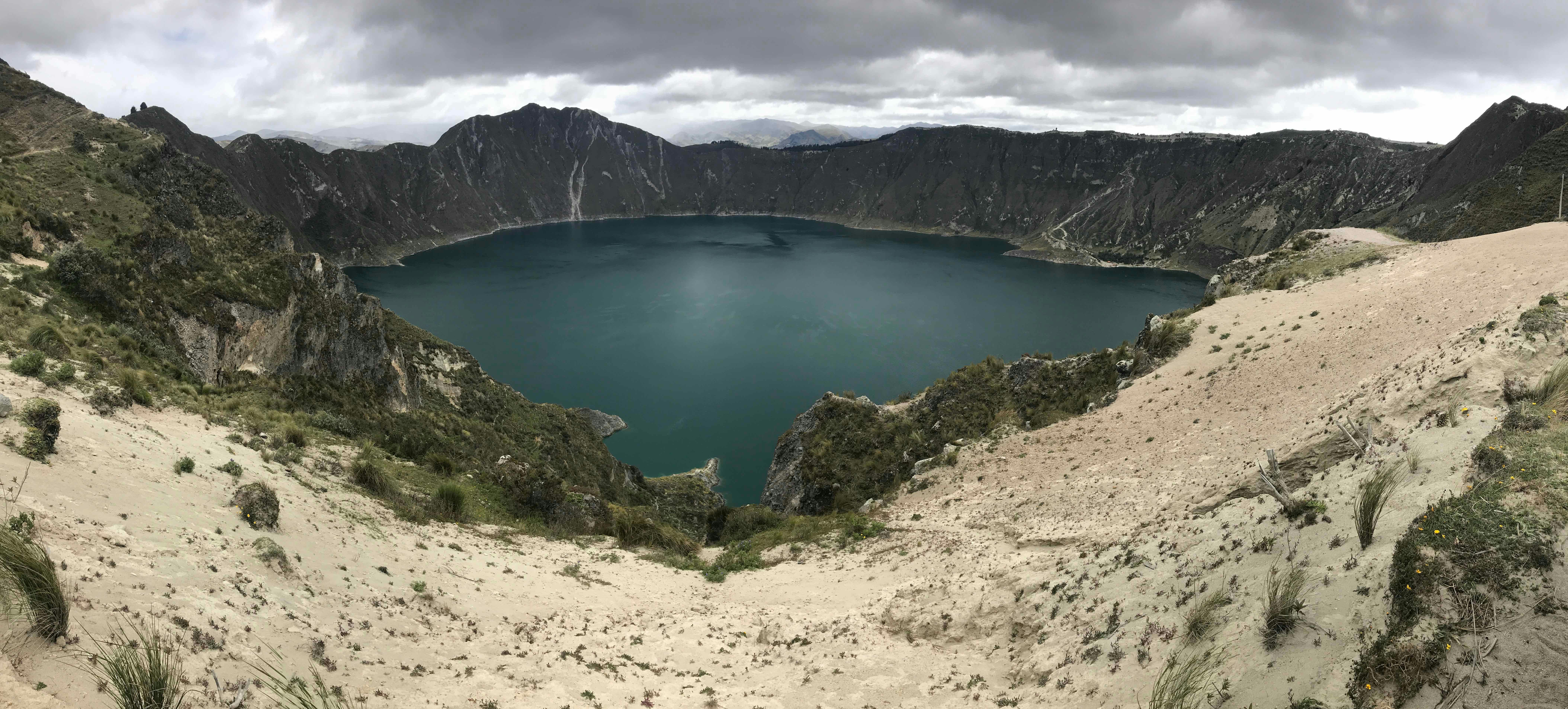 A panoramic view of Quilotoa Lake, showcasing its vibrant turquoise waters surrounded by steep volcanic cliffs under a dramatic sky.