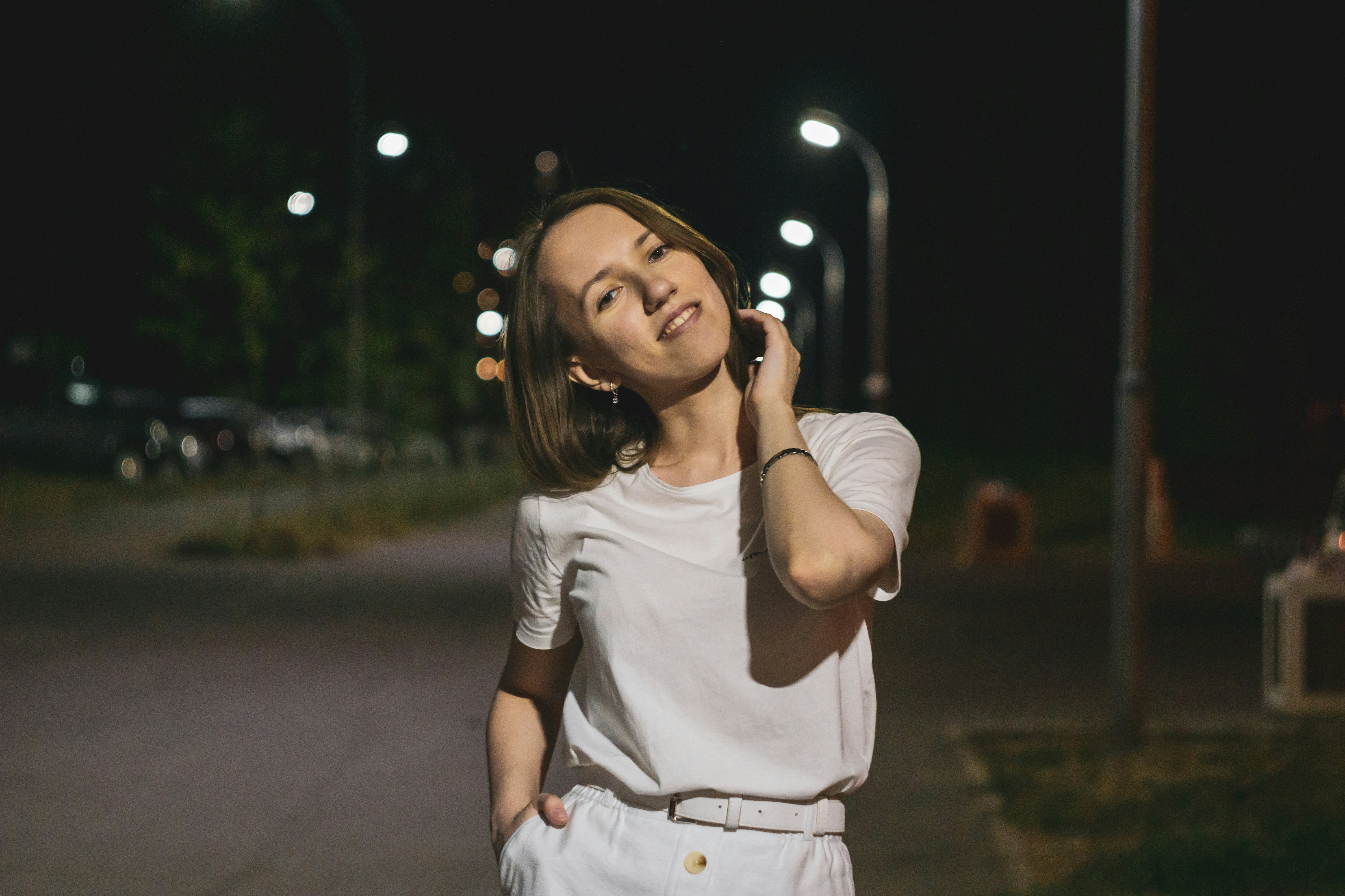 Woman in a white t-shirt standing on an illuminated street at night, casually posing with one hand on her neck.
