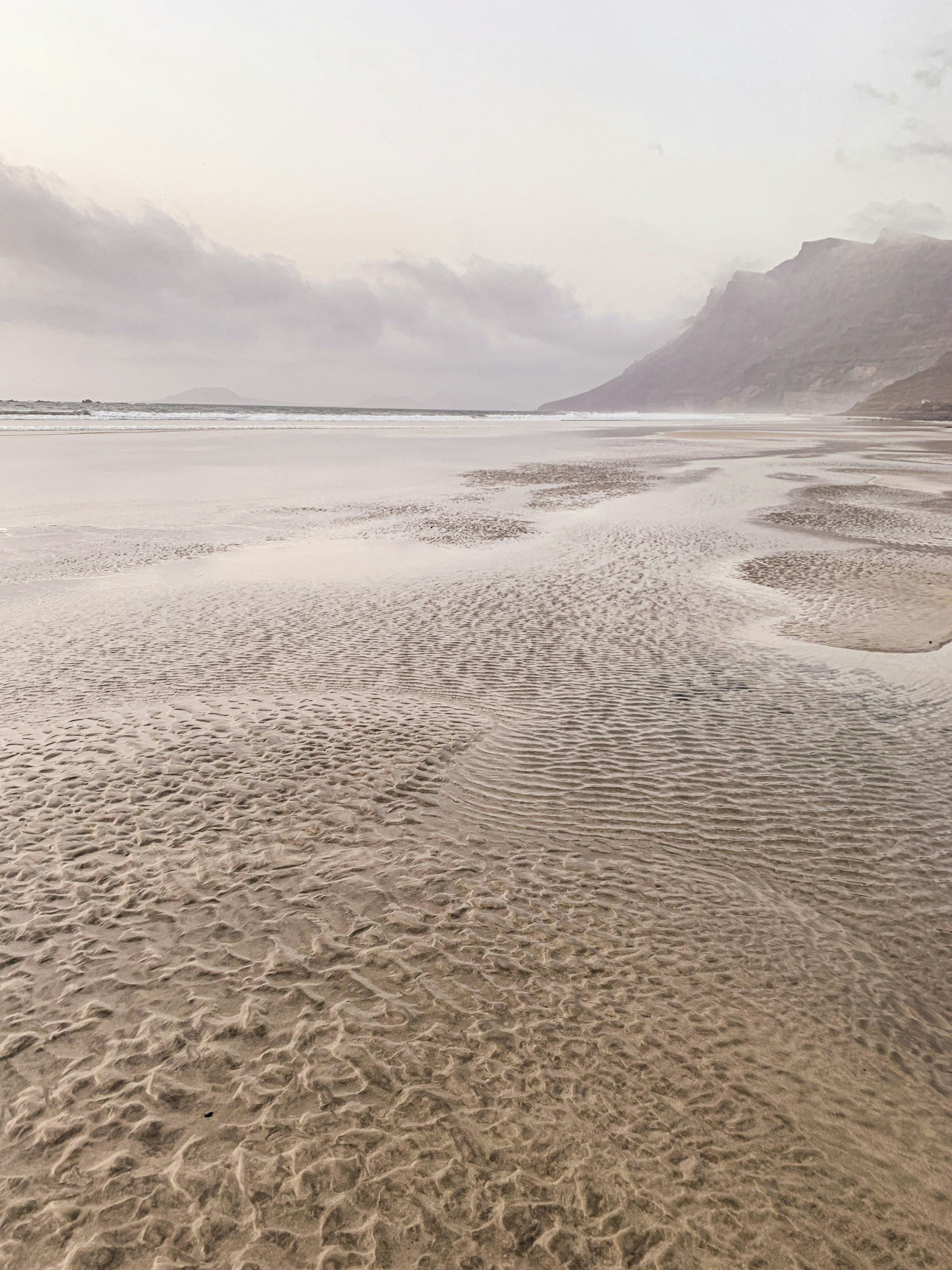 Gentle waves lapping against a textured sandy beach under a moody sky. The scene captures the serene interaction between land and sea.