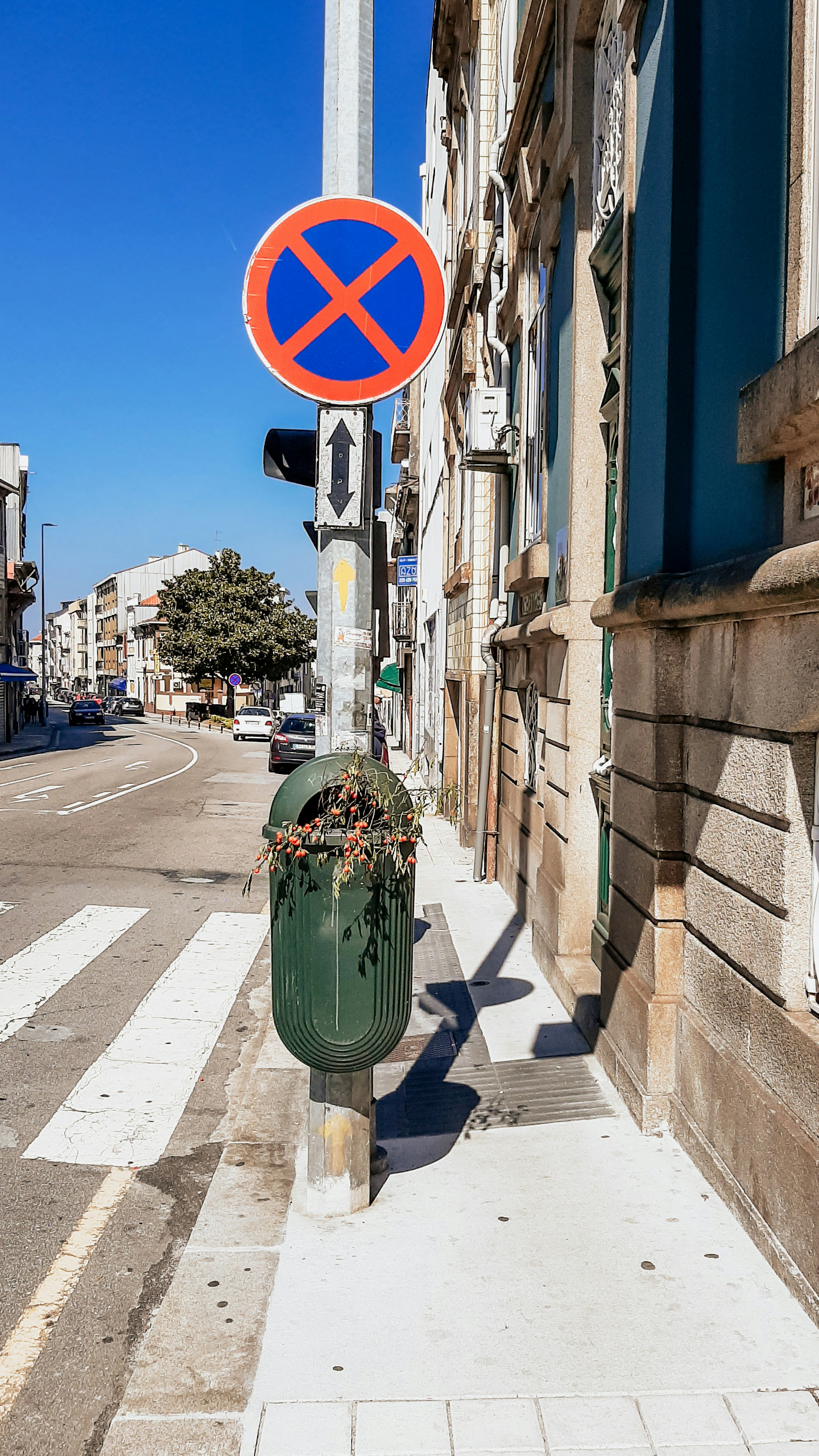 Sunlit city street with a tall no-parking sign and a leaning green trash can beside historic stone buildings and a zebra crossing.