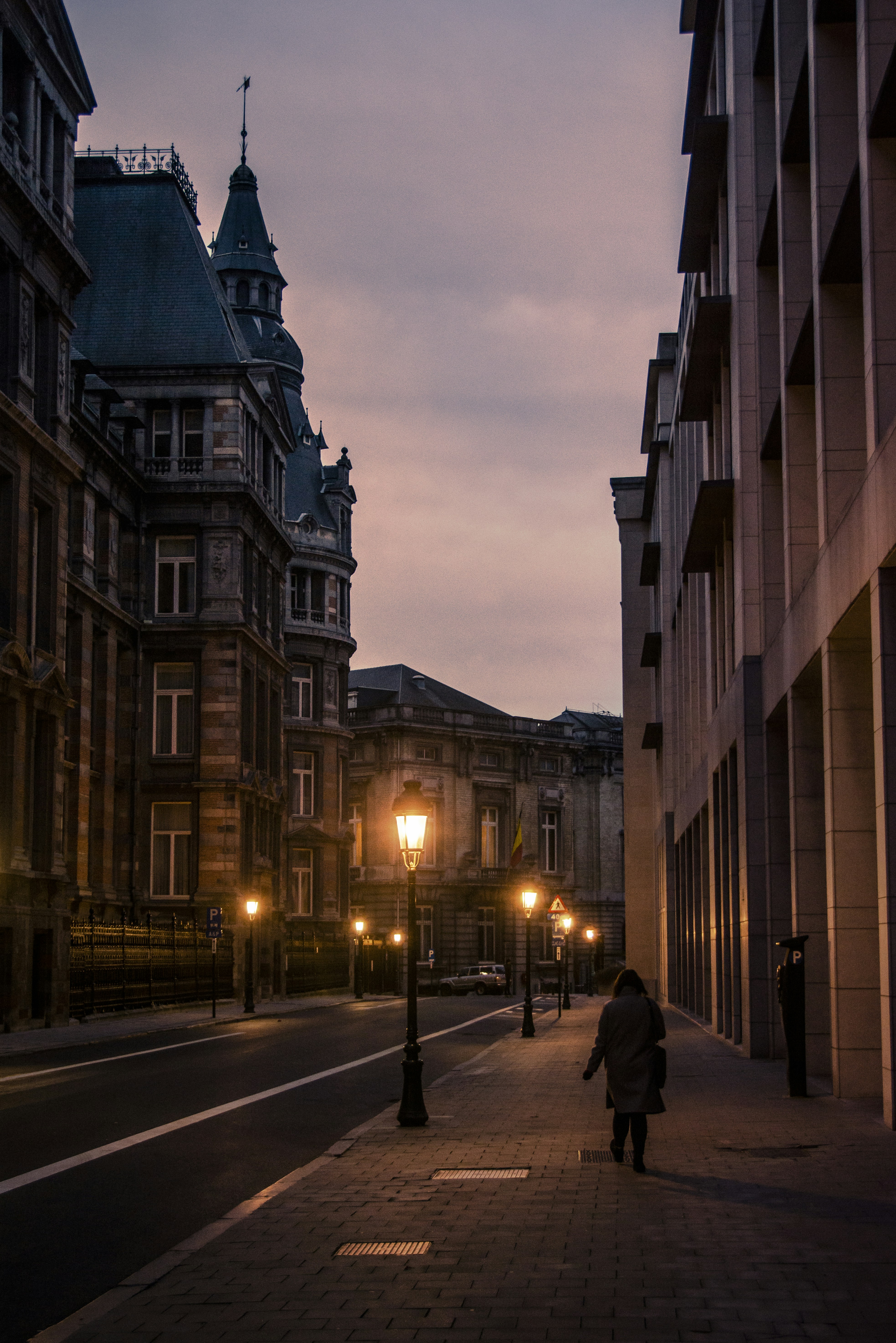 A solitary figure walks along a cobblestone street flanked by historic buildings and modern architecture, illuminated by vintage street lamps. The soft twilight sky casts a serene ambiance.