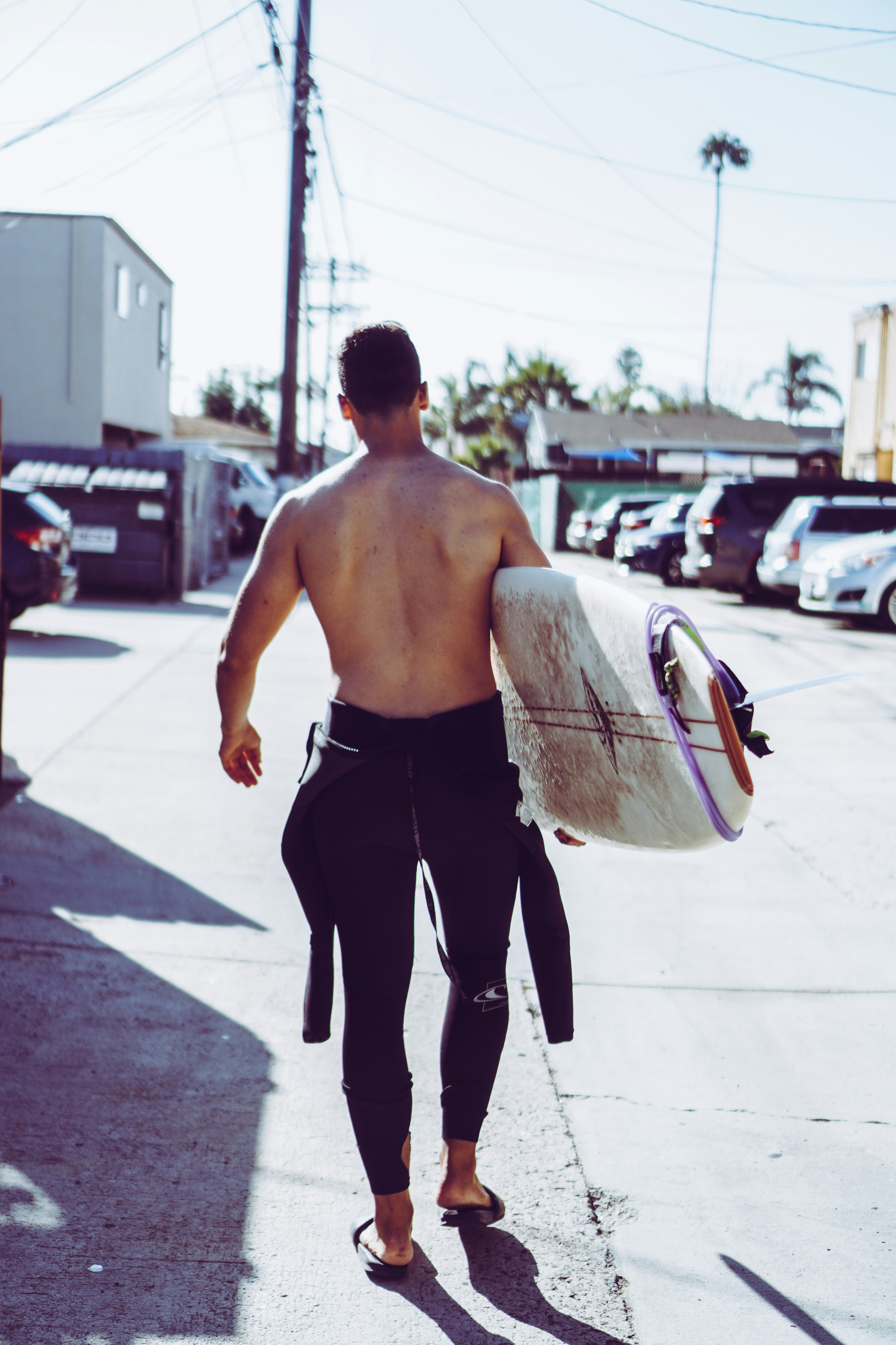 Sunlit Curbside Surf: A Shirtless Surfer Hauls His Board Down a Sunny Street