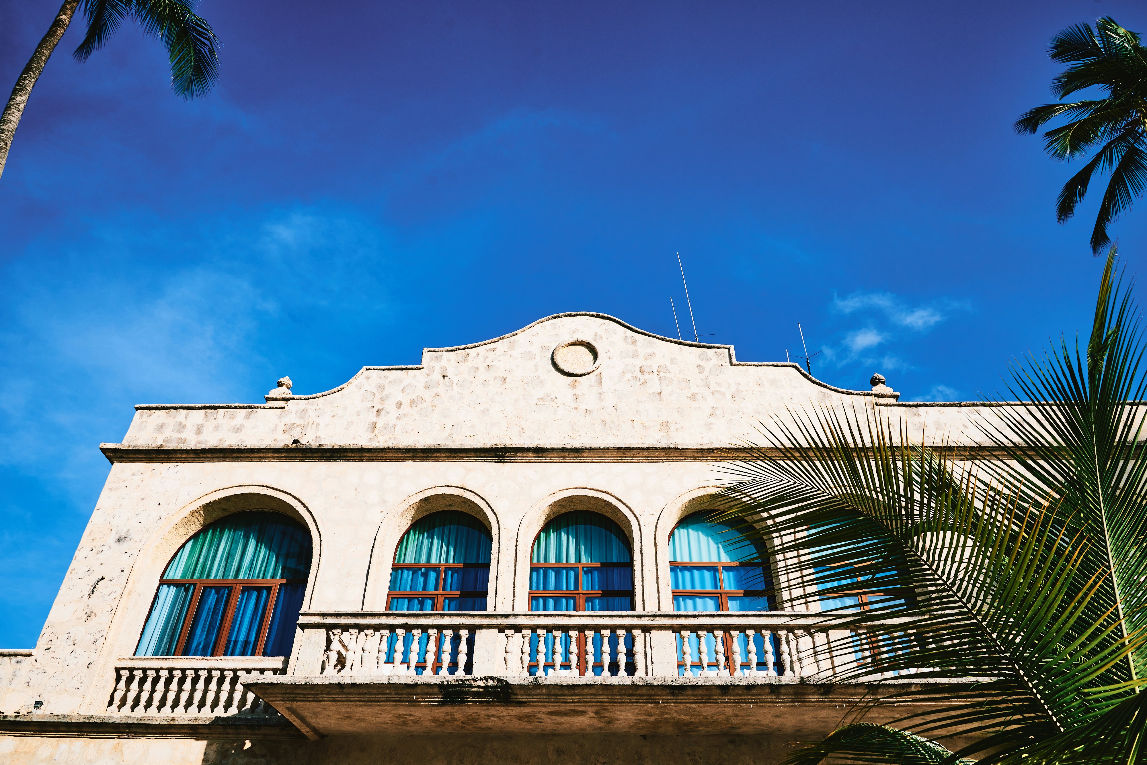 Historic building with arched windows and vibrant blue curtains, framed by palm leaves against a clear sky.