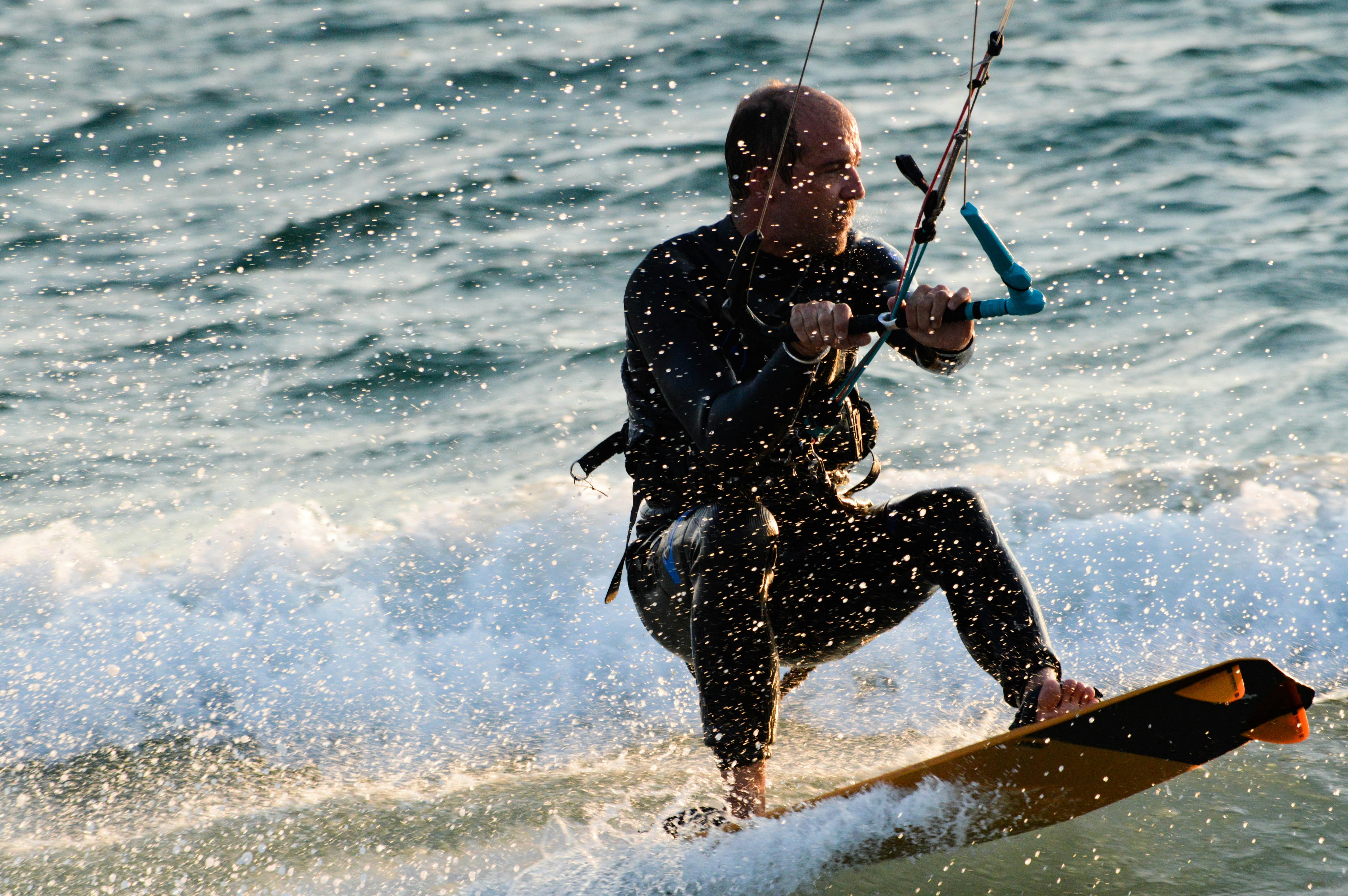 Kiteboarder in black wetsuit carving through waves on an orange board under bright daylight.