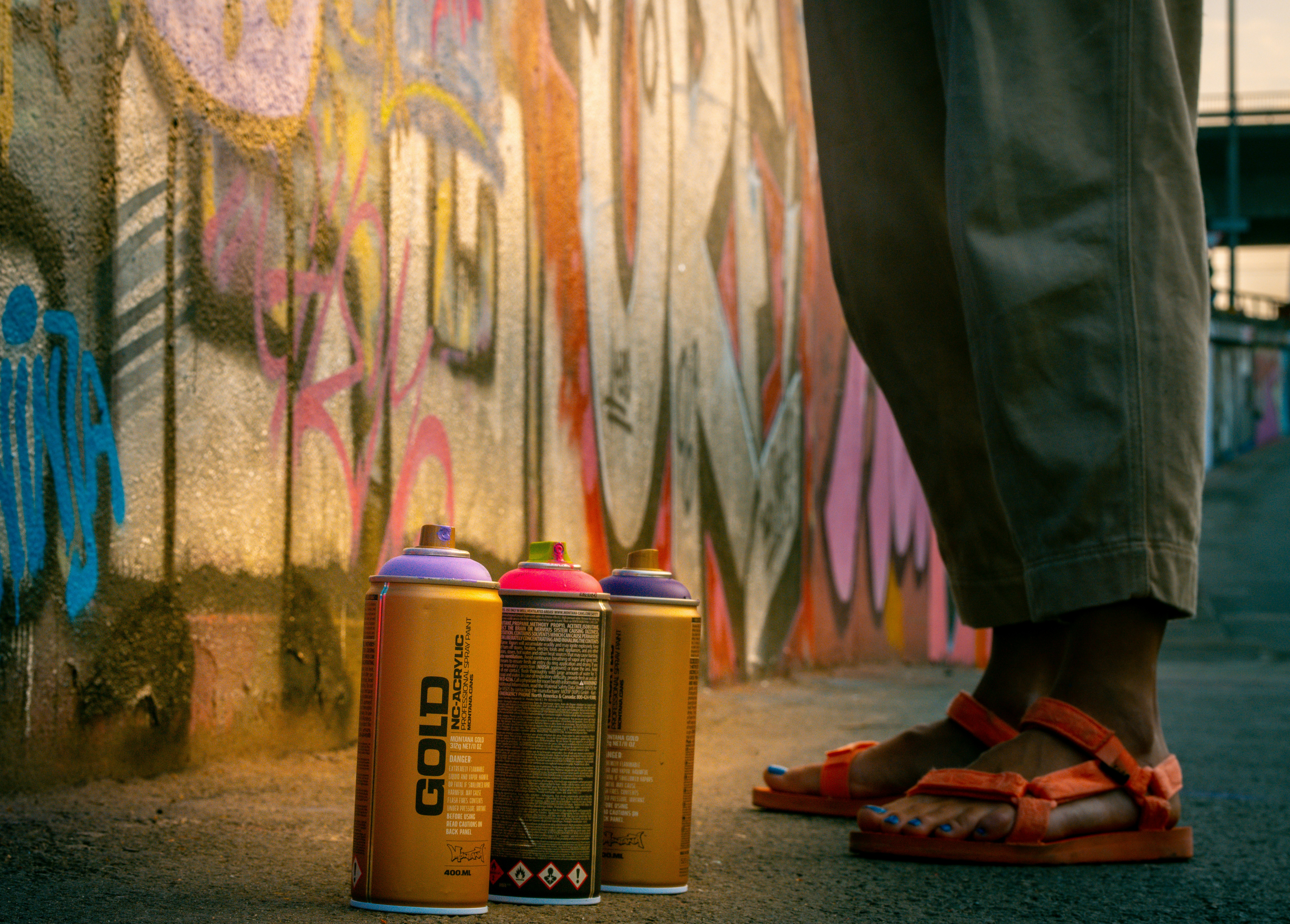 Three spray paint cans stand on the ground beside a person wearing orange sandals, with a vibrant graffiti wall in the background.